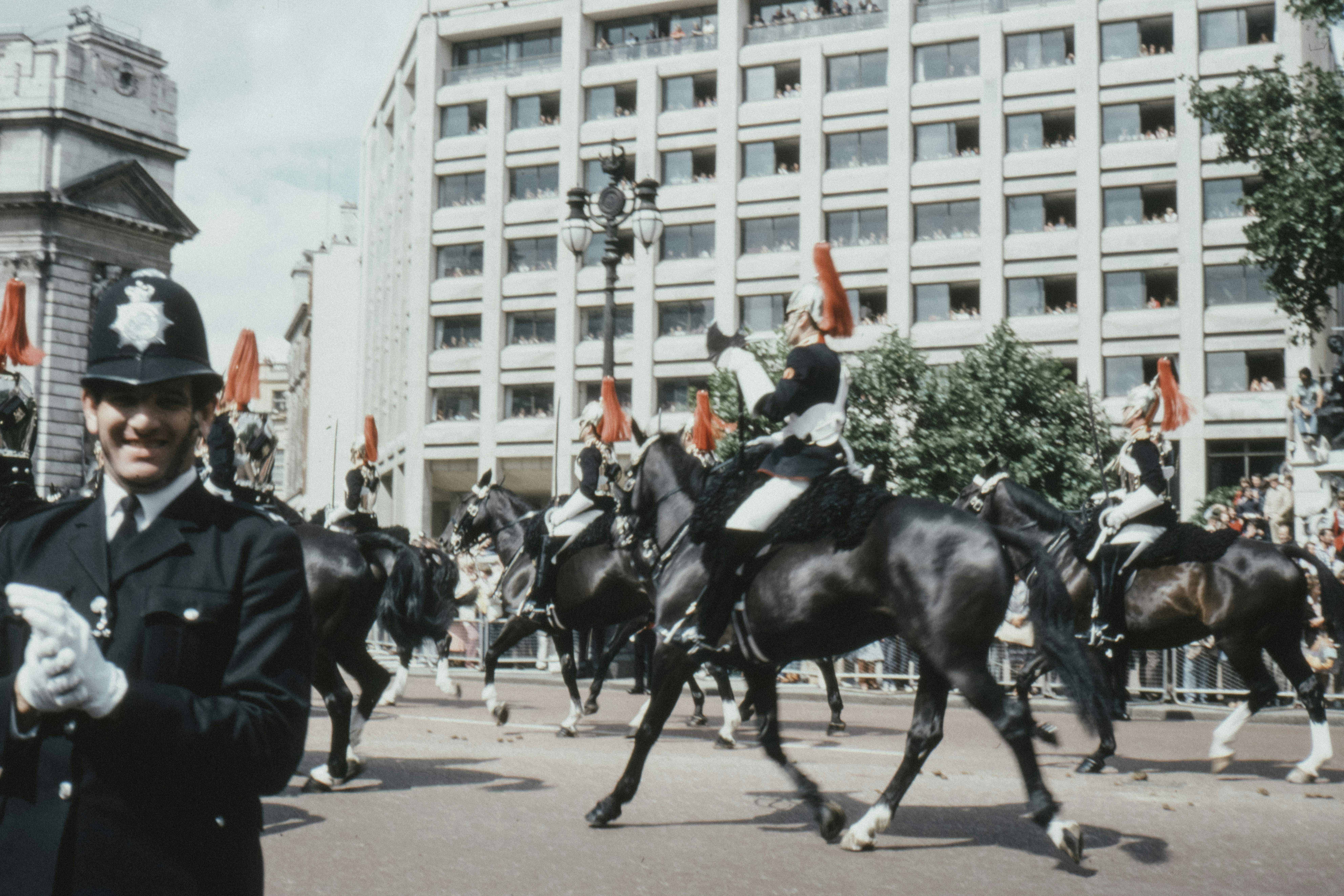 Color photograph of a police officer in the foreground while a line of mounted cavalry with red plumes marches through a city square, with a tall modern building in the background.