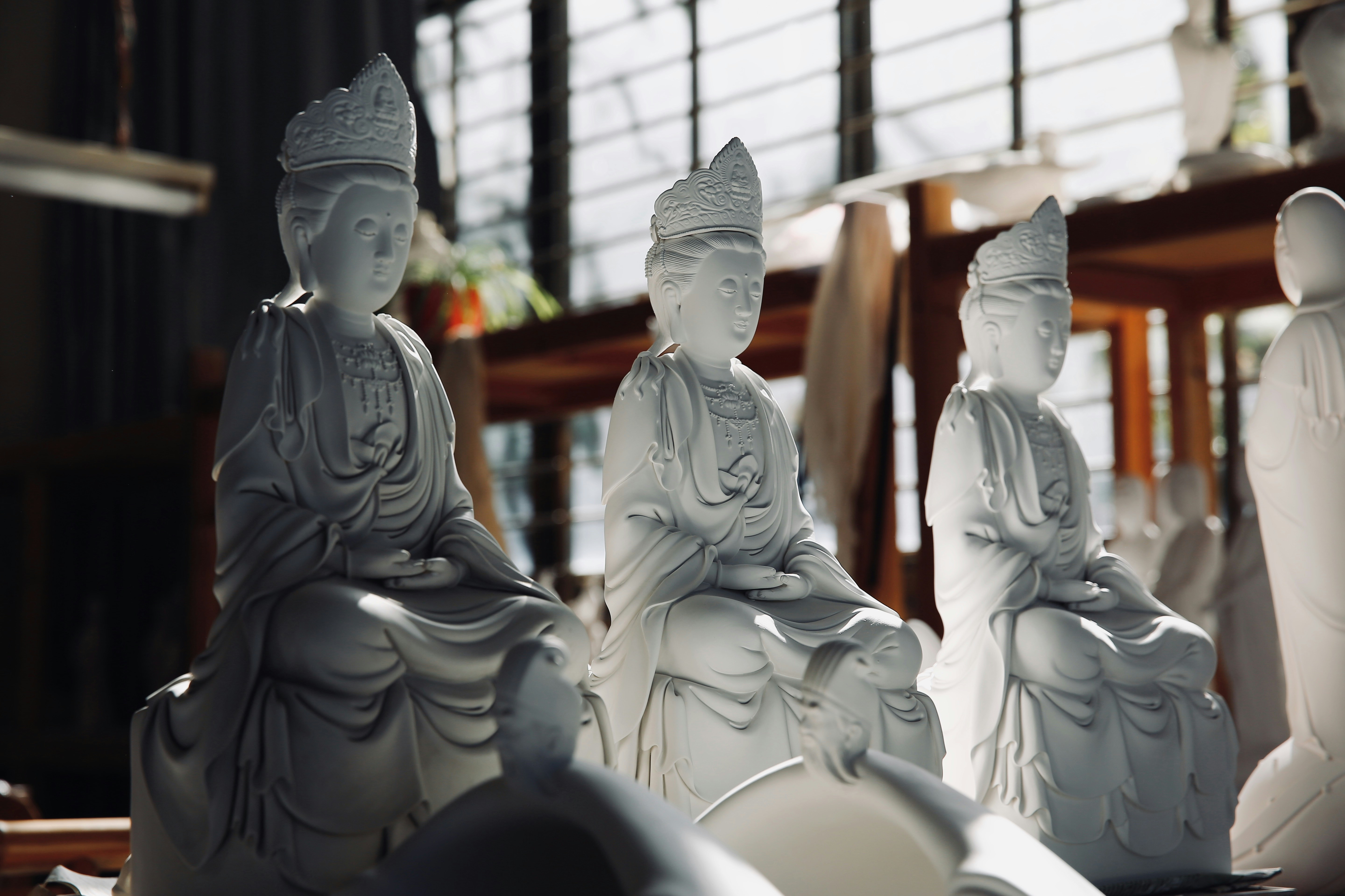 Three intricately crafted porcelain statues of seated Buddhas, illuminated by soft natural light filtering through a workshop window.