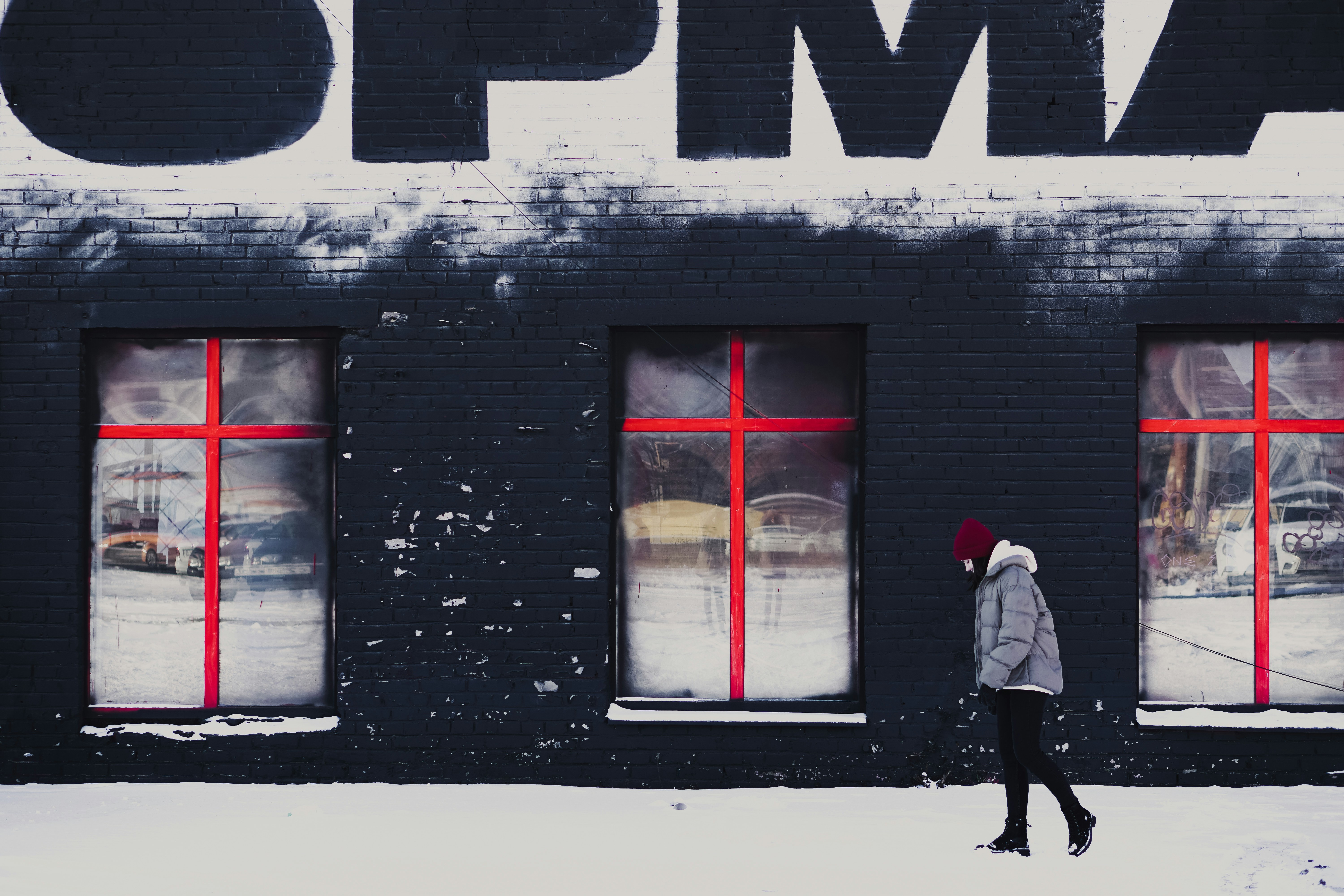 A person walks past a stark black wall adorned with red cross-patterned windows, surrounded by a blanket of snow.