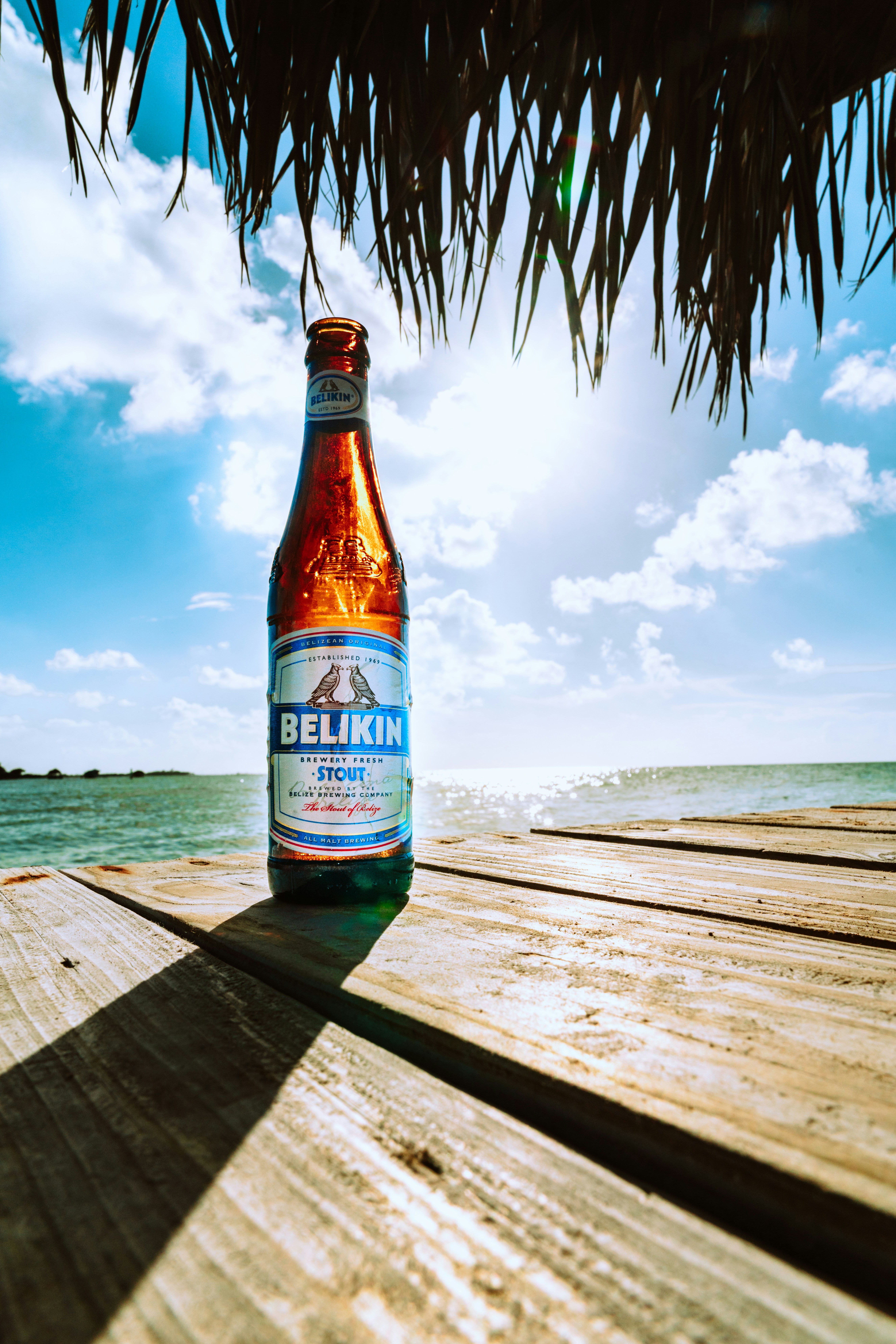 A Belikin Stout bottle stands on a wooden deck, with the sun shining brightly in the background and a serene ocean view beyond.