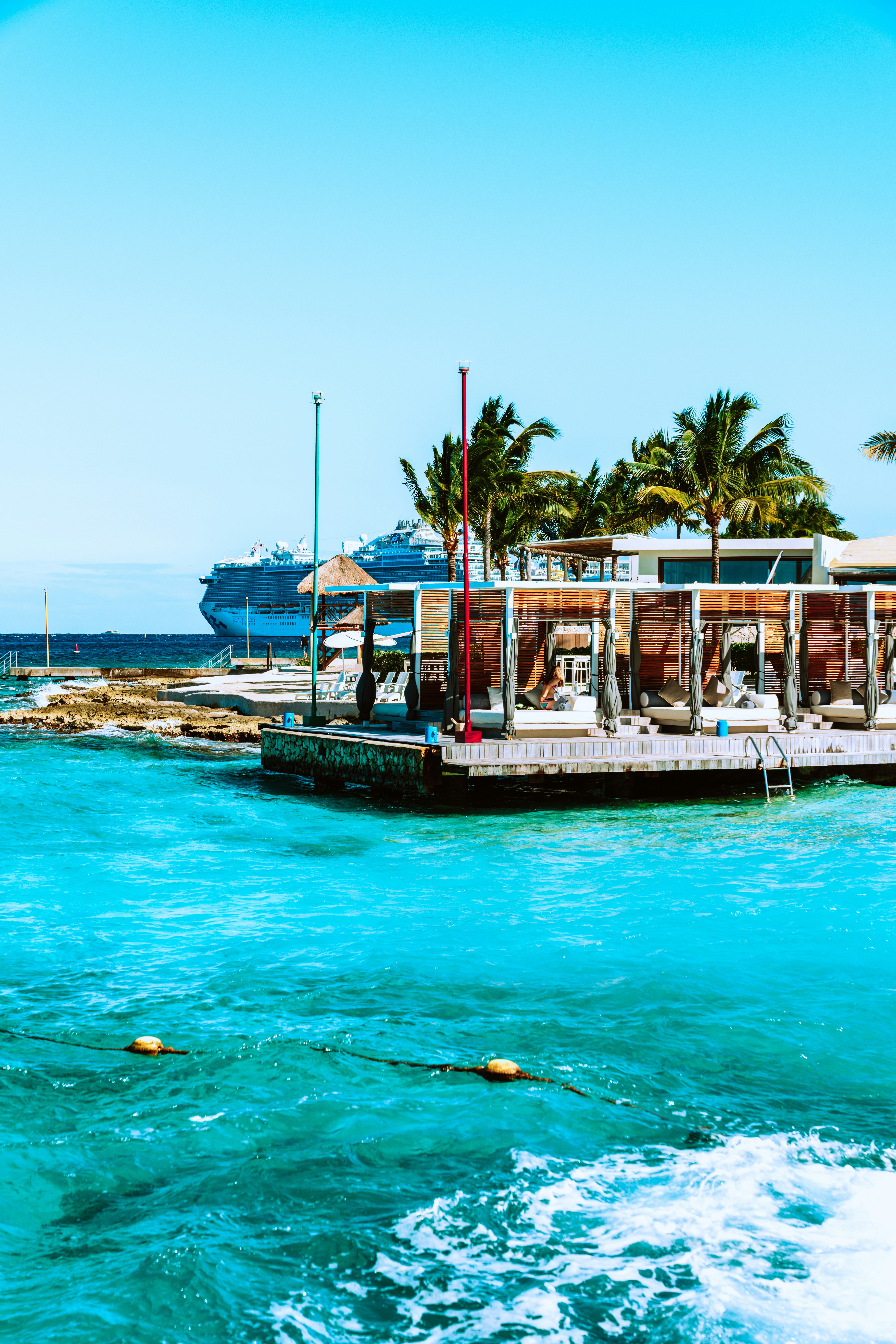 Private cabanas line the shore against a backdrop of turquoise ocean and distant cruise ships.