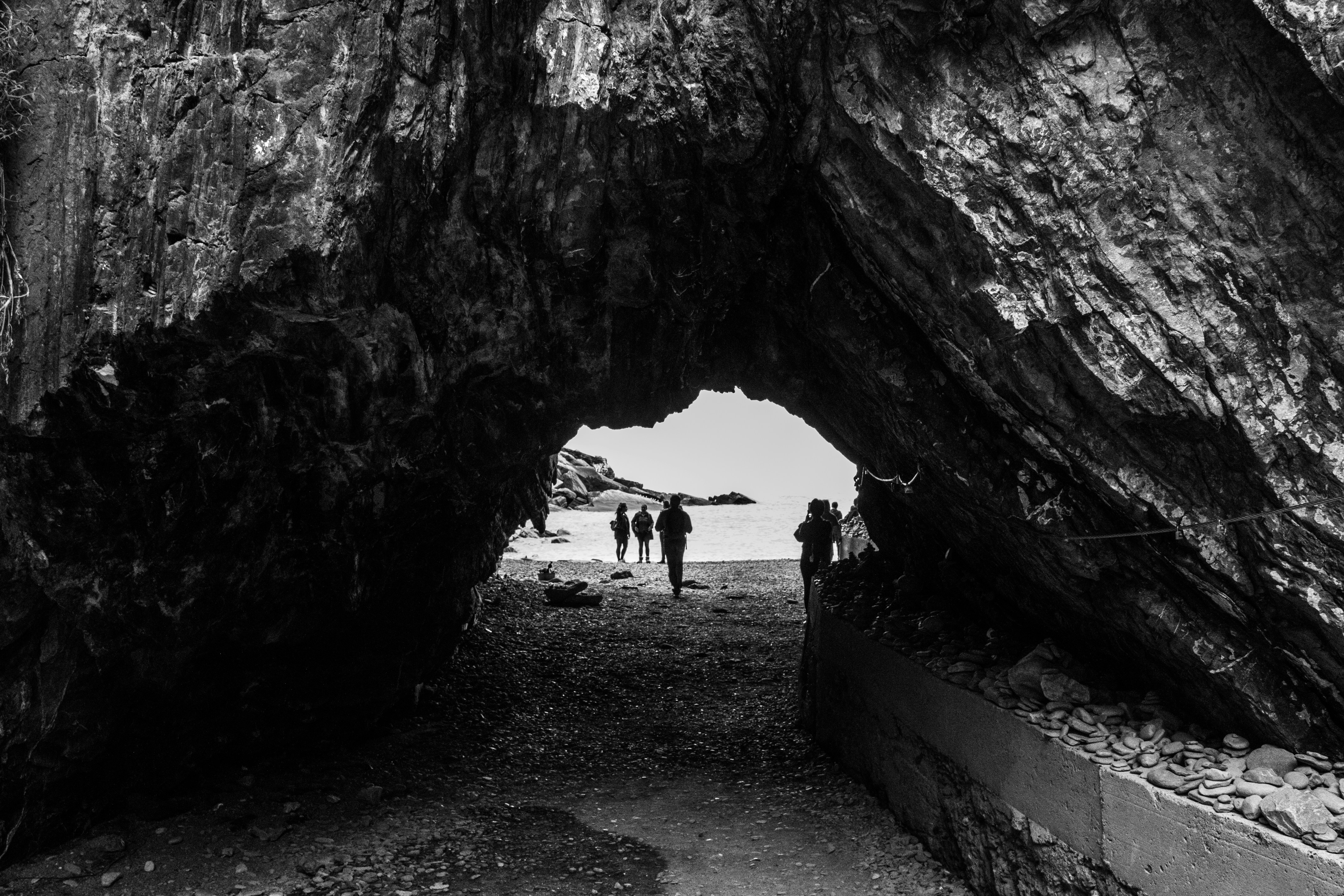 Silhouetted figures walking through a rocky cave entrance with a view of the beach and sea beyond.