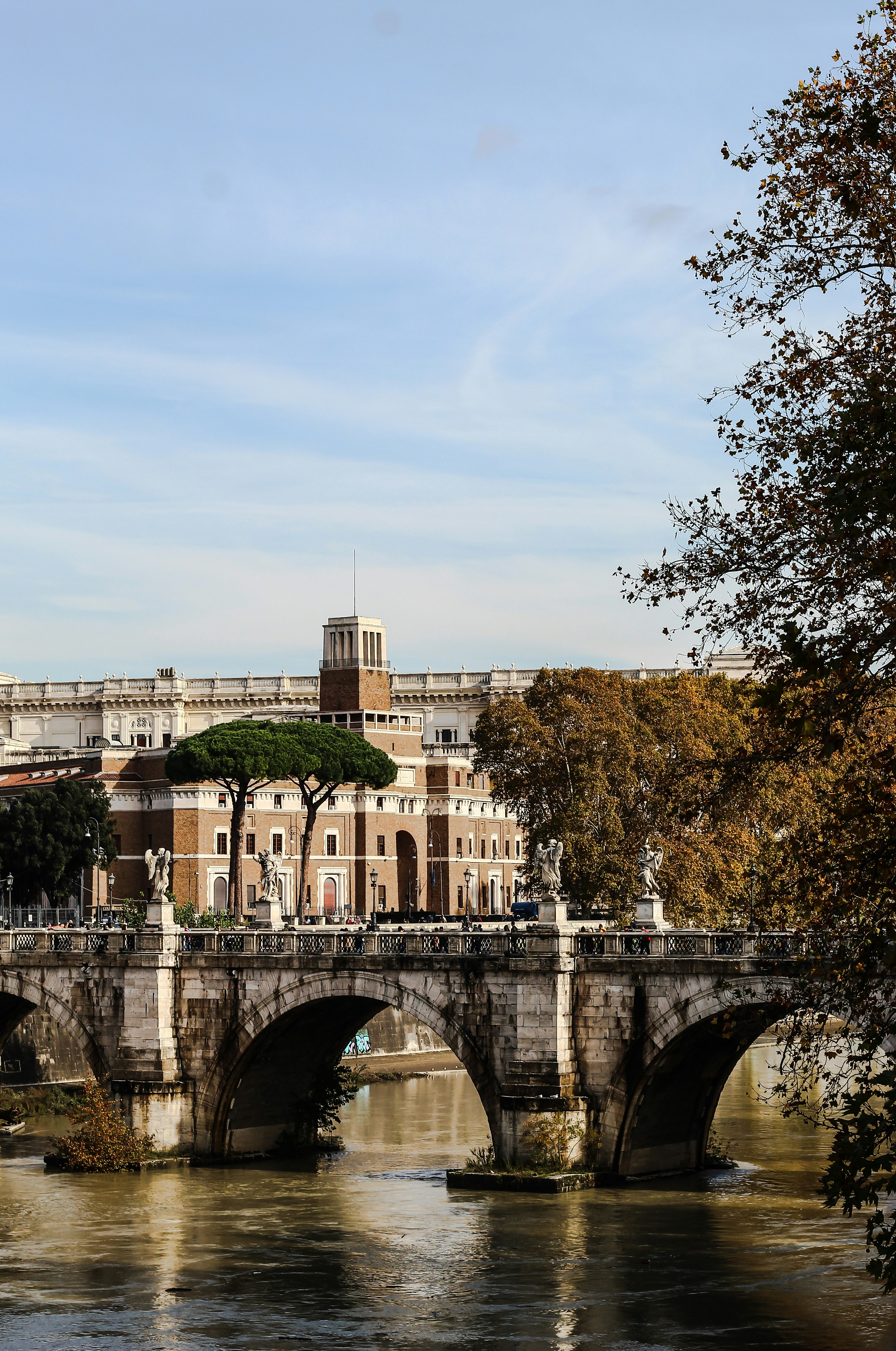 Historic stone bridge adorned with statues, leading to a grand building surrounded by autumn foliage. The serene river reflects the architectural beauty.