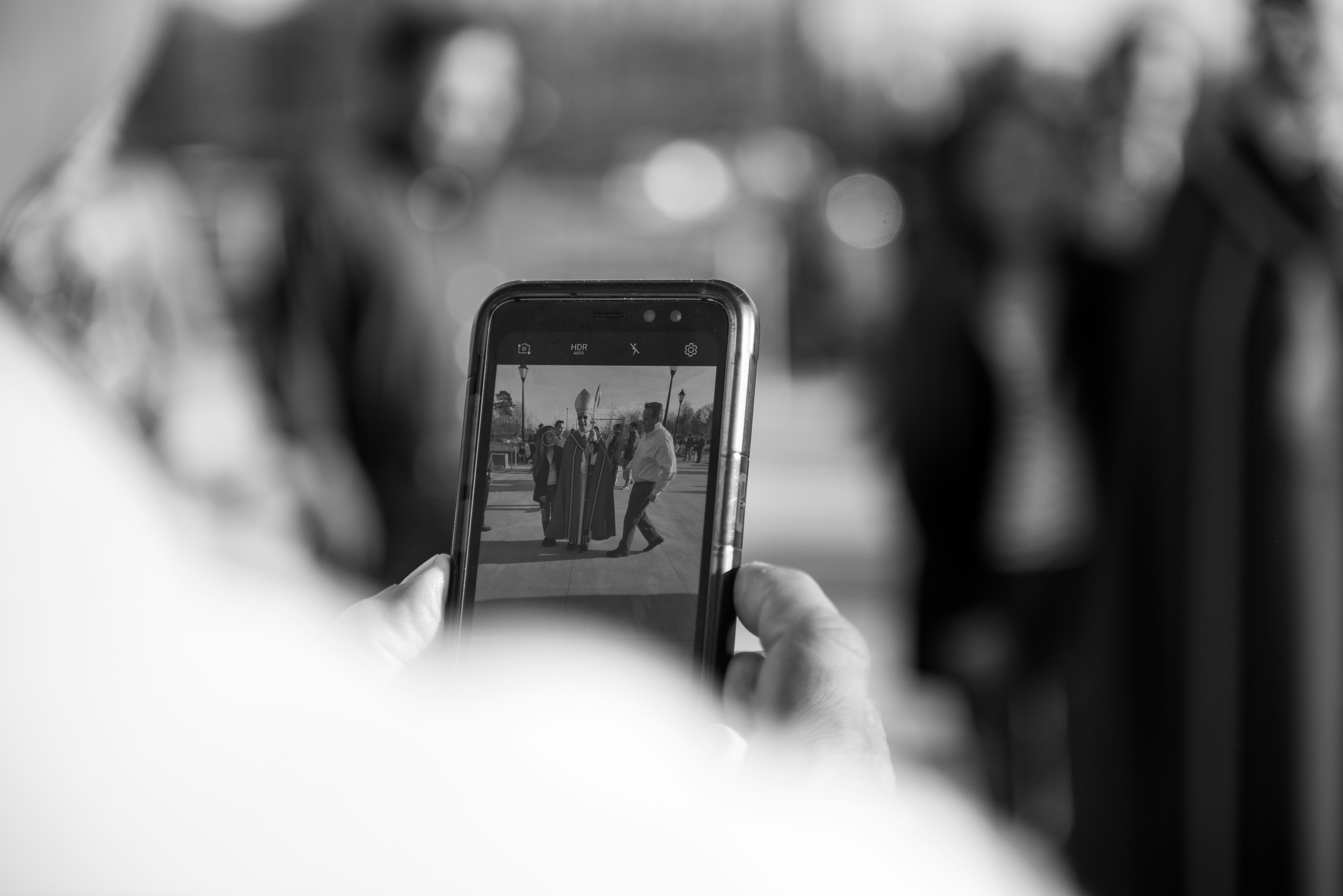 Black and white photo of a person taking a picture with a smartphone, focused on a group of people in the background.