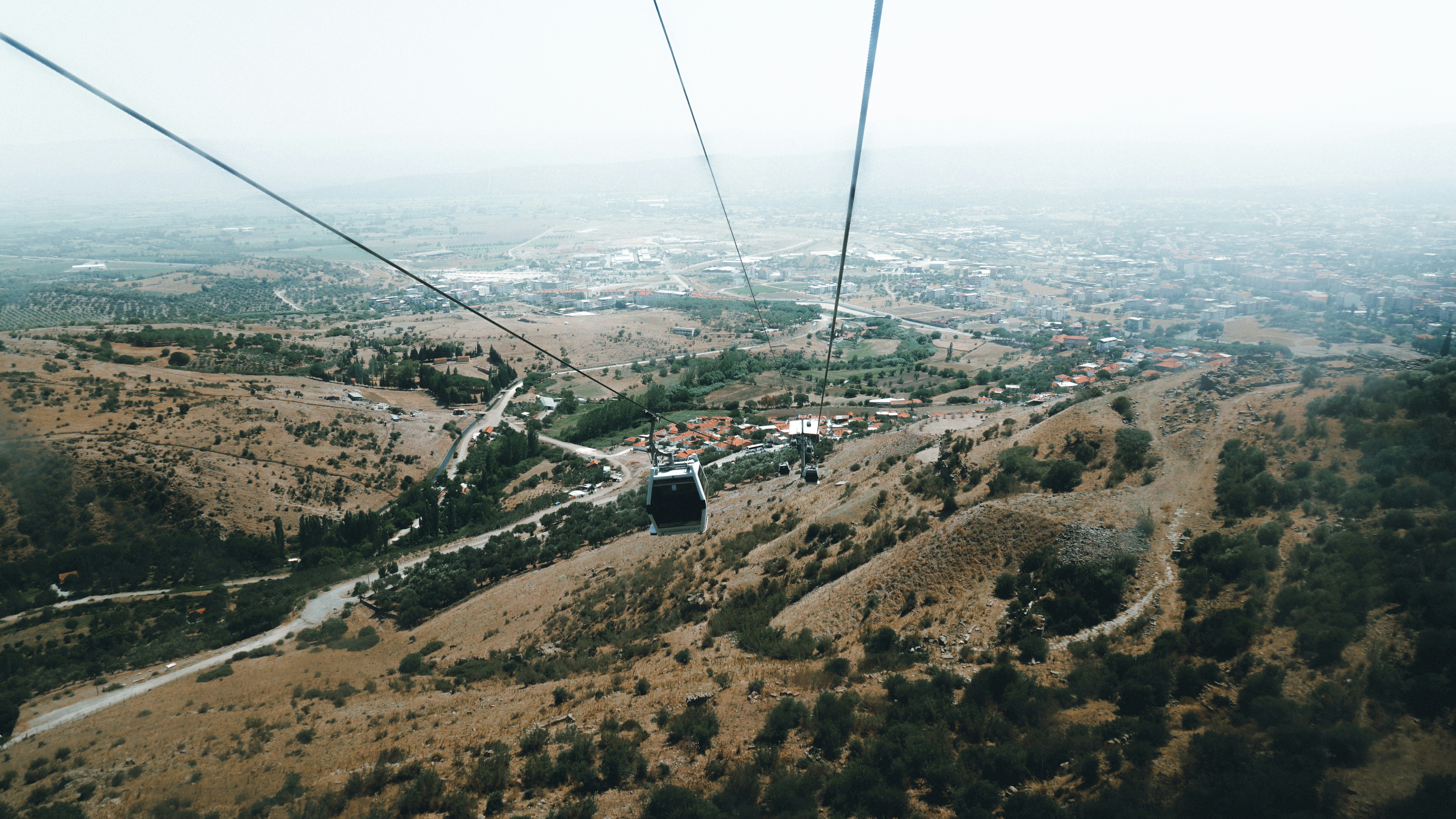 Cable cars traverse above a sprawling landscape of arid hills and distant cityscape under a hazy sky.