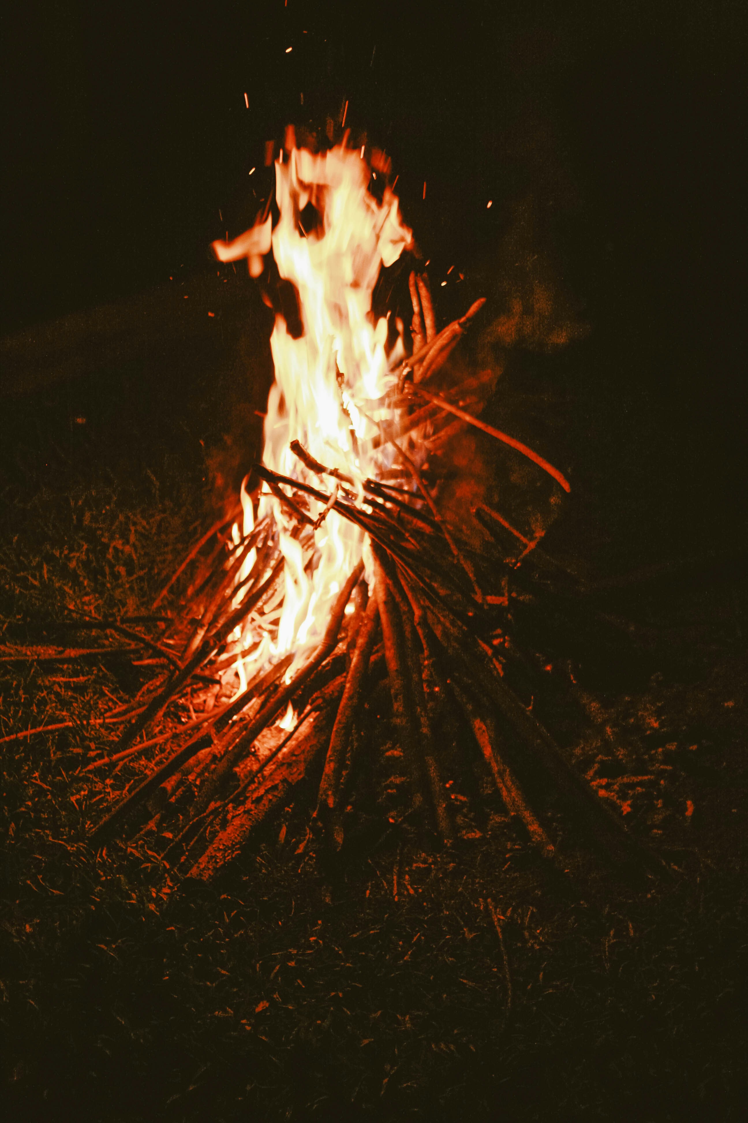 A vibrant campfire crackling amidst a dark backdrop, with wooden sticks arranged in a tepee formation. The flames flicker, illuminating the surrounding grass.