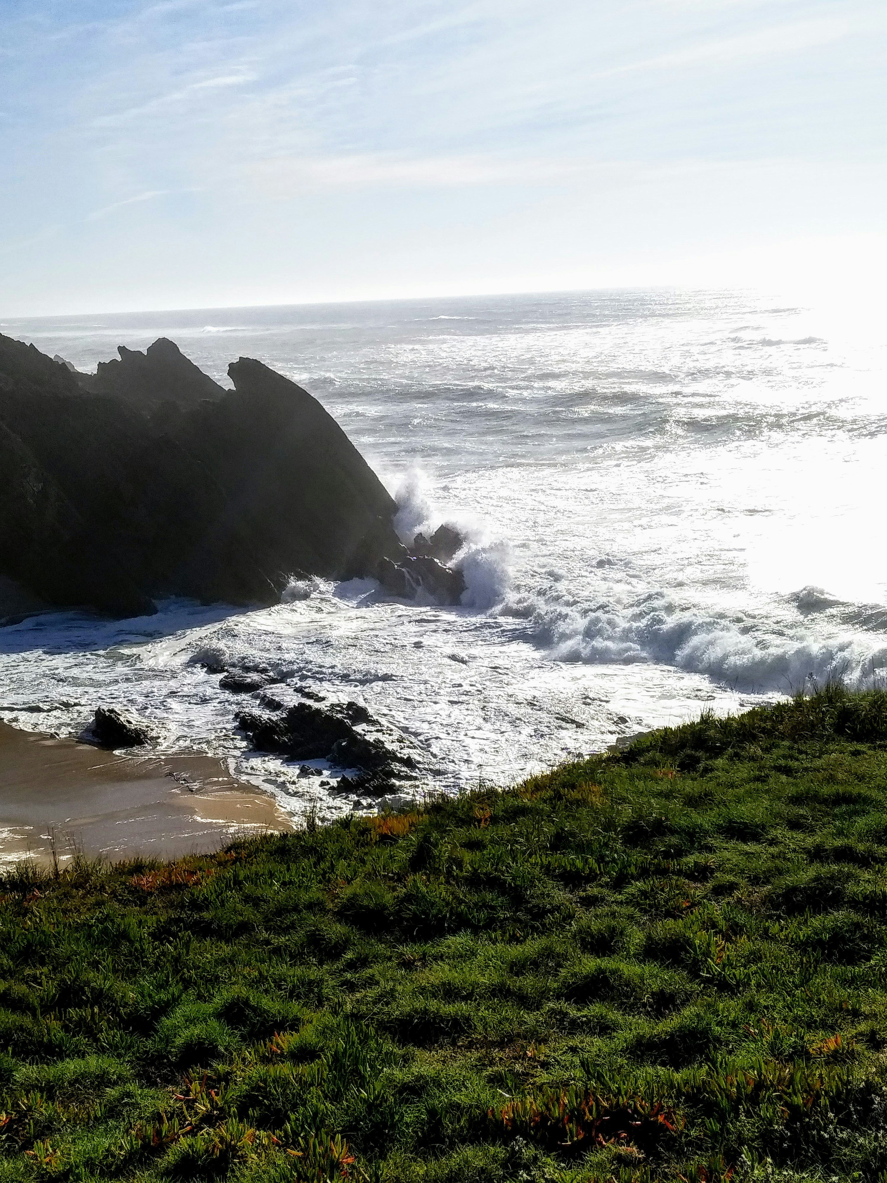 Rugged coastline with crashing waves against dark rocks, framed by lush green vegetation. A serene coastal scene under a bright sky.