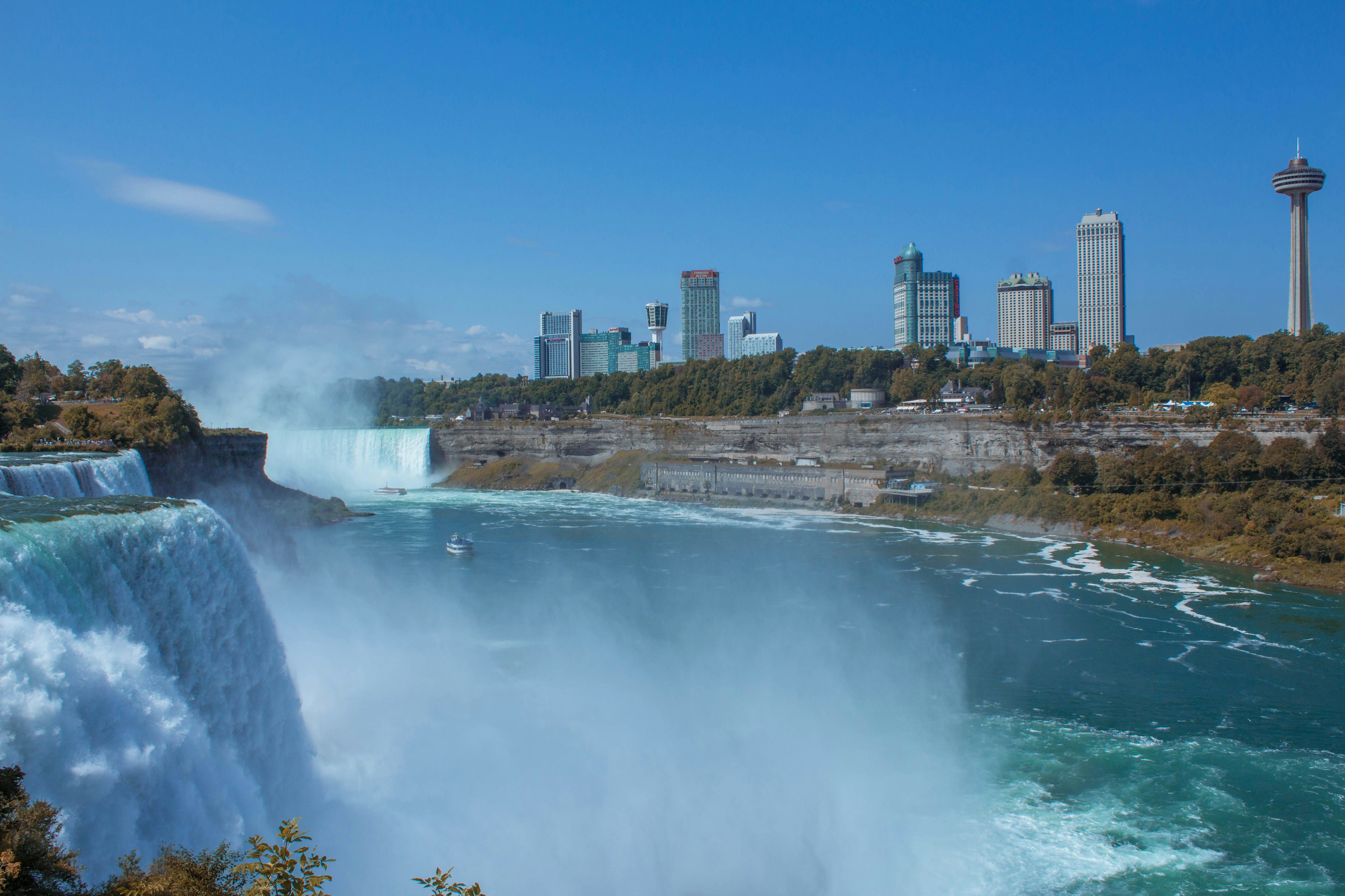 Majestic waterfalls cascade into a vibrant river, framed by a bustling city skyline and clear blue skies.