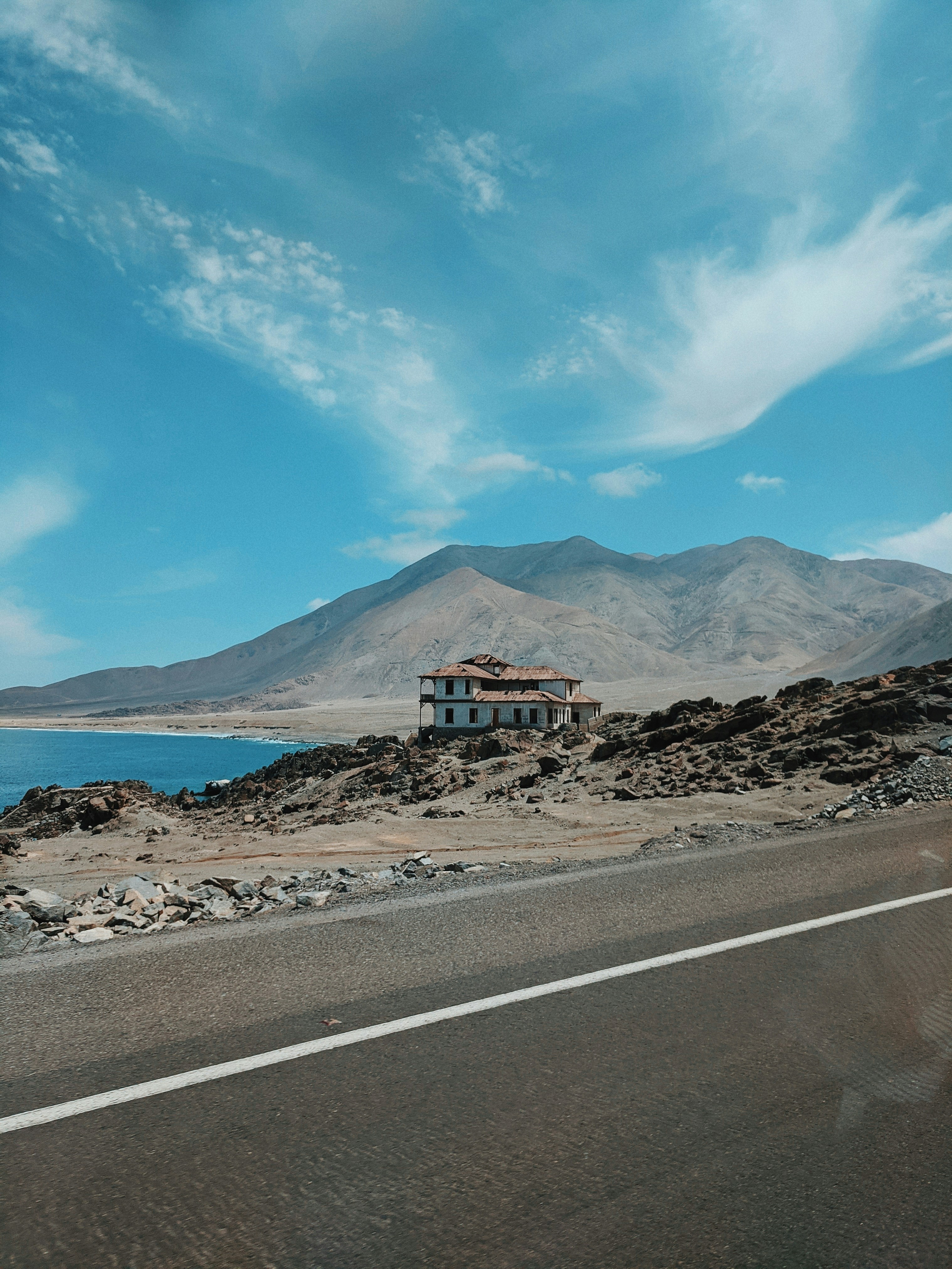 Abandoned house perched on rocky coastline with mountains in the background under a bright blue sky.
