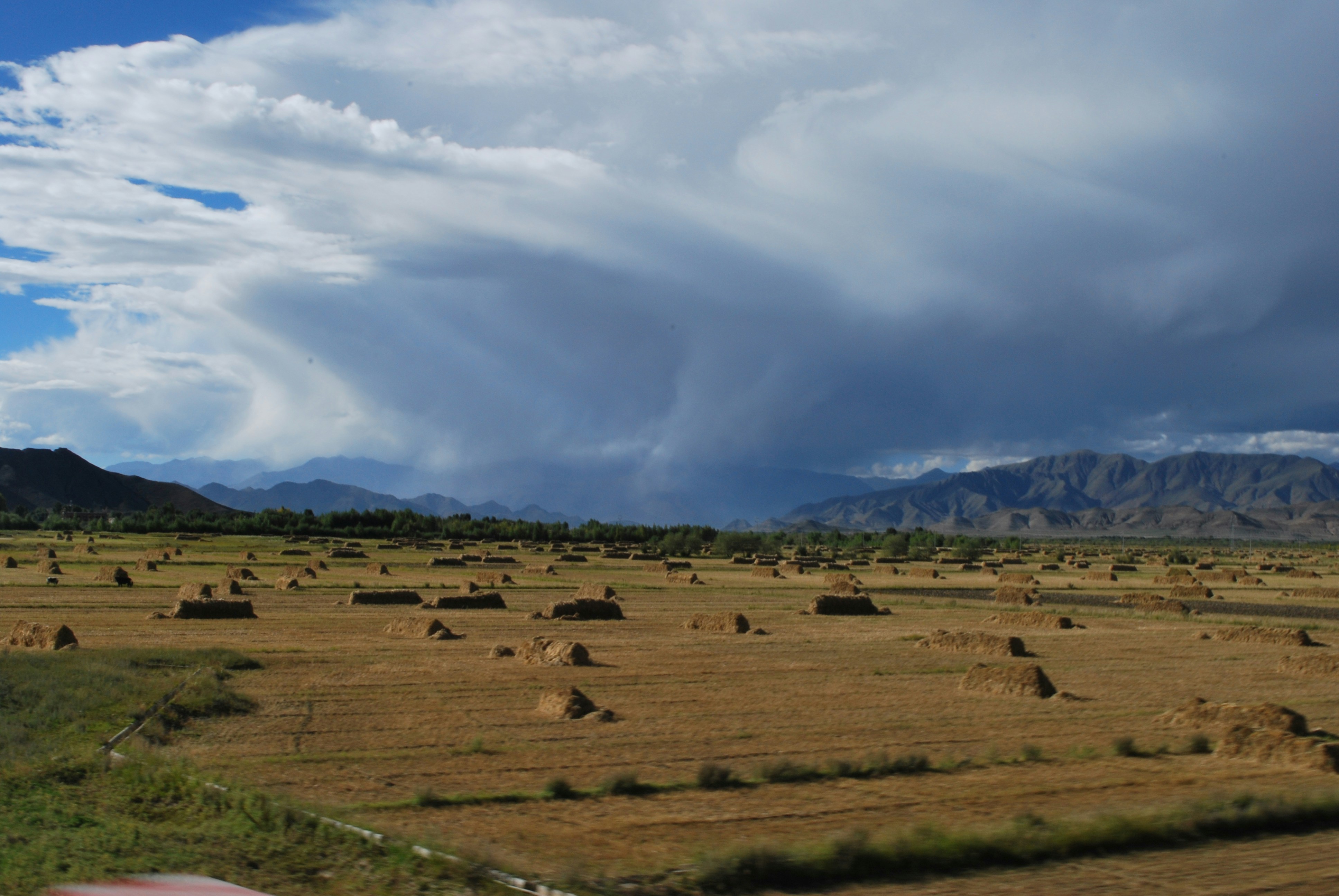 Vast agricultural landscape with haystacks under dramatic cloud formations and distant mountains. A blend of earth and sky showcases nature's vastness.