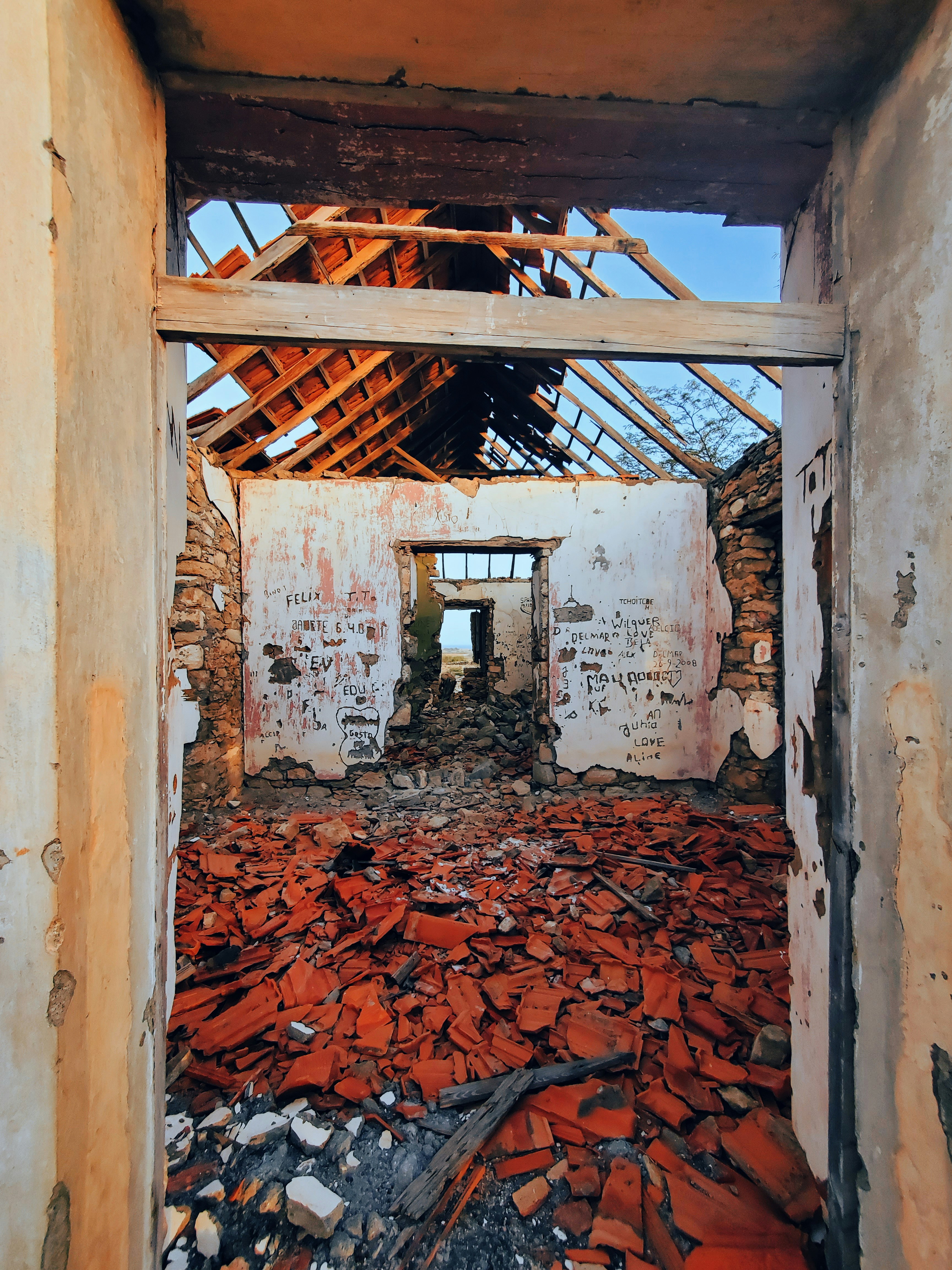 Interior of a dilapidated building with scattered red tiles and peeling walls, revealing remnants of past writings. Sunlight filters through the broken roof, highlighting the decay.