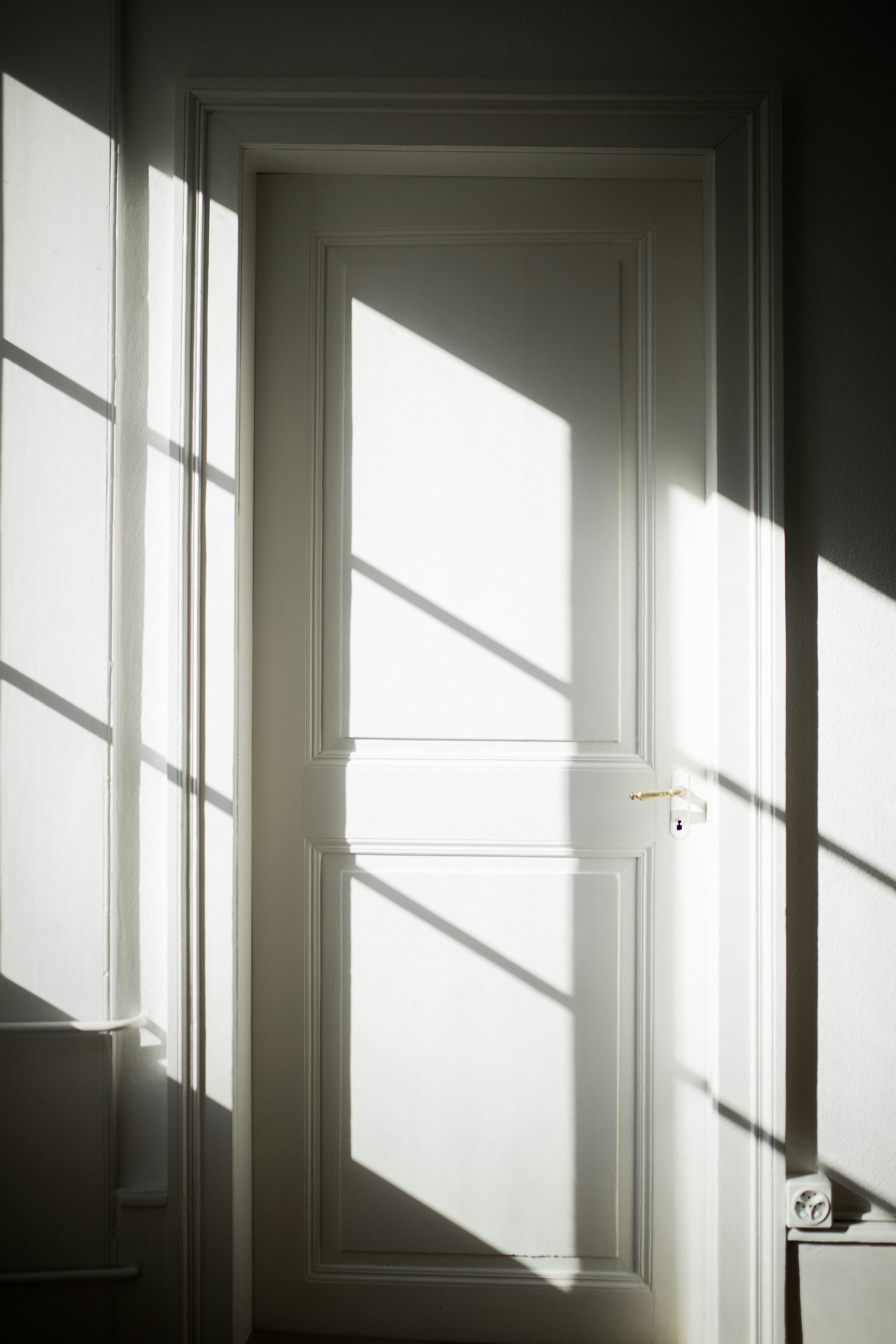 Sunlight streams through a doorway, casting intricate shadows on the wall. The simplicity of the white door contrasts with the dynamic patterns of light.
