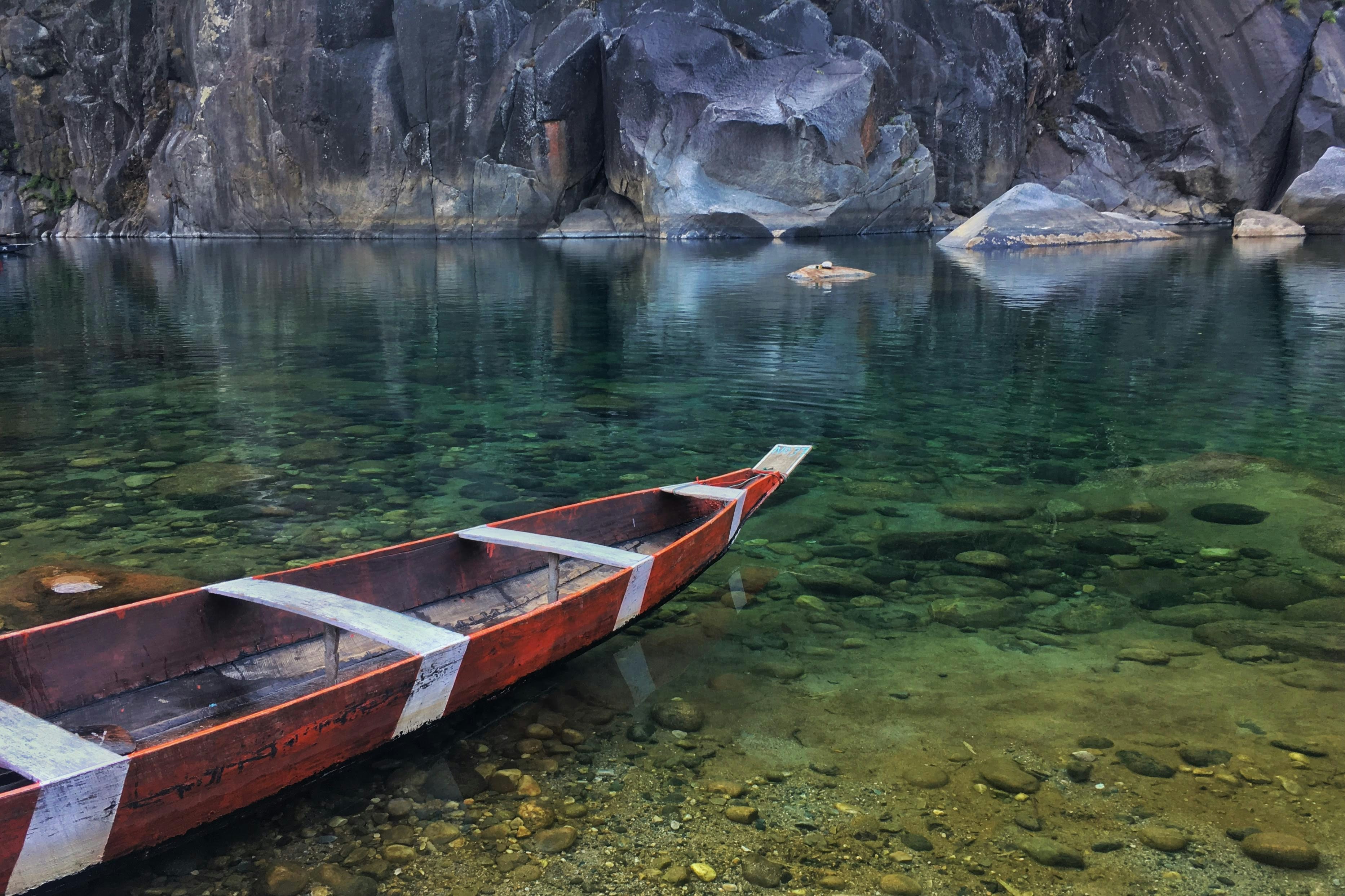 A weathered wooden canoe rests quietly on the clear, tranquil waters, surrounded by rocky cliffs and smooth stones beneath the surface.