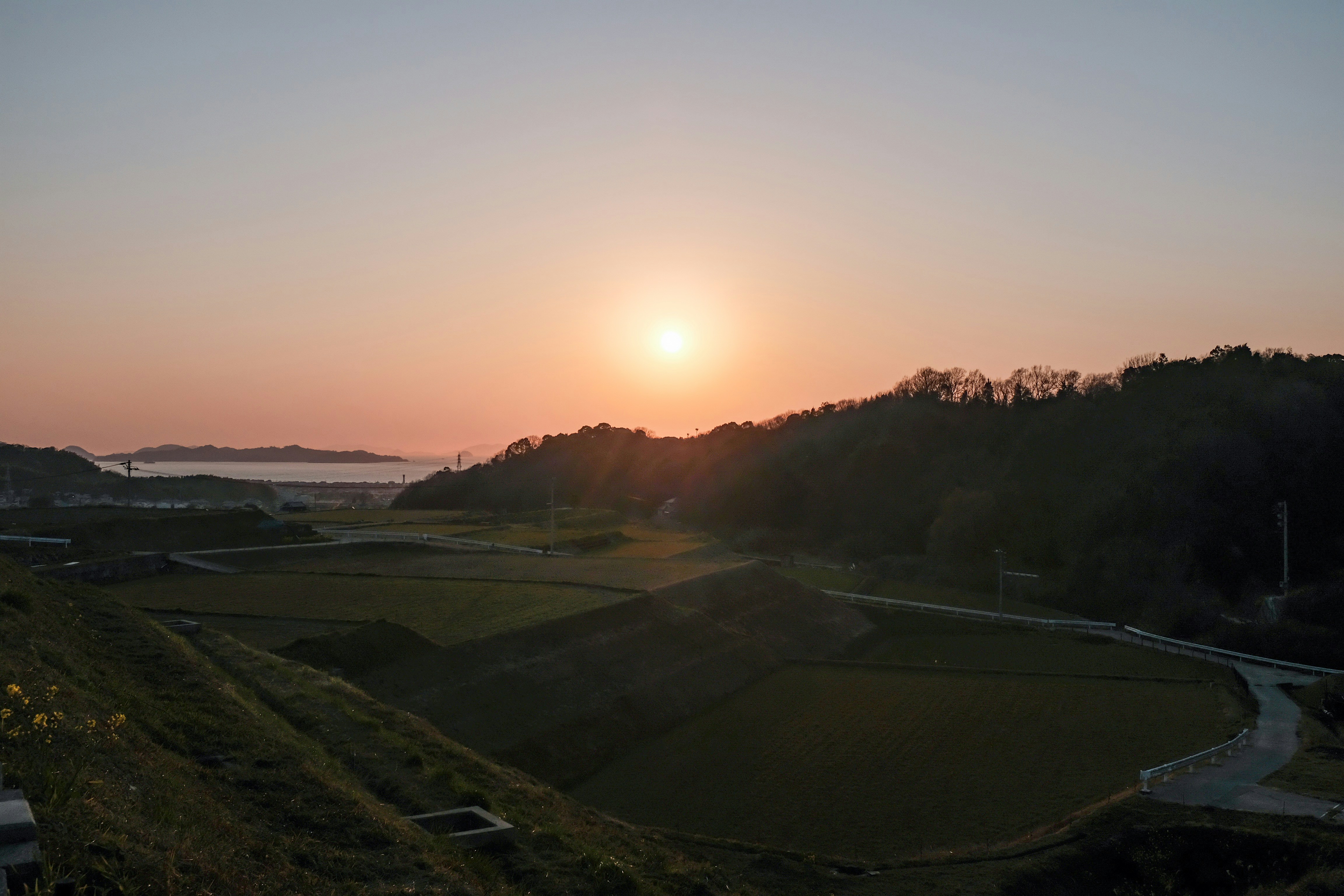 Golden sun setting over lush terraced fields, casting a warm glow across the landscape. The horizon blends soft hues of orange and pink.