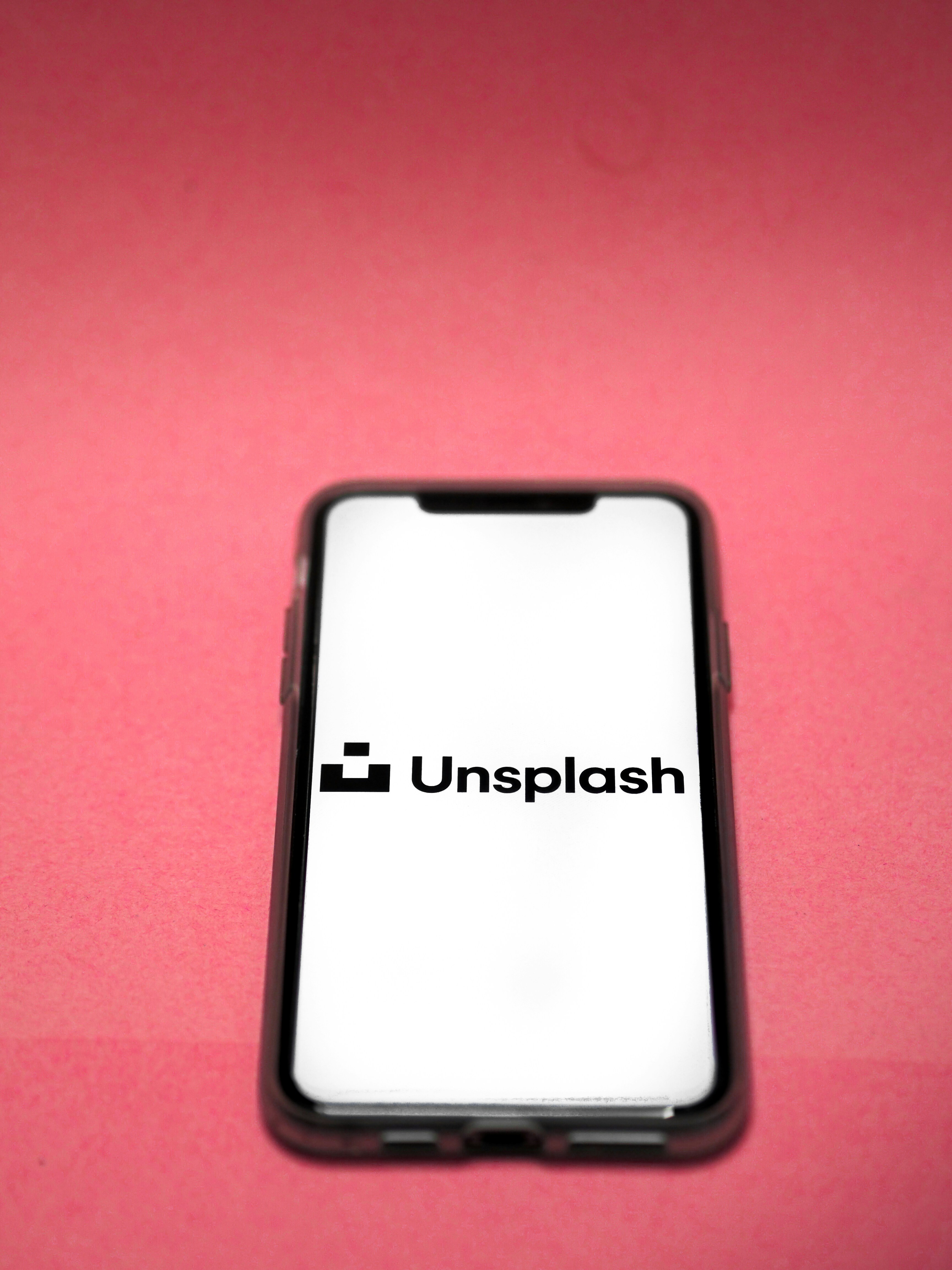 Smartphone foreground displays the Unsplash logo on a vivid pink backdrop. The setup emphasizes branding against a minimal, color-rich background.