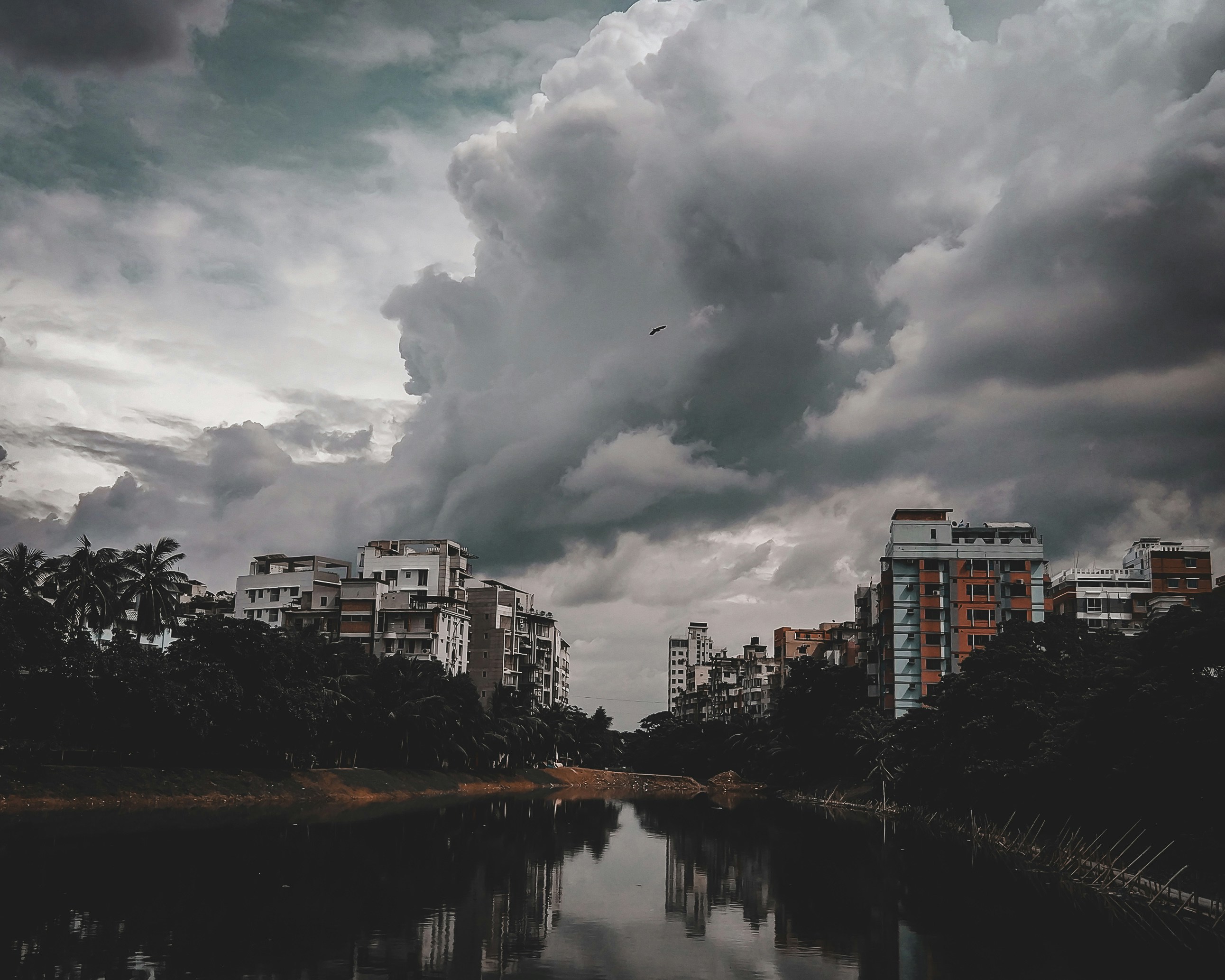 Dark clouds loom over a tranquil river, reflecting the surrounding urban landscape and lush greenery.
