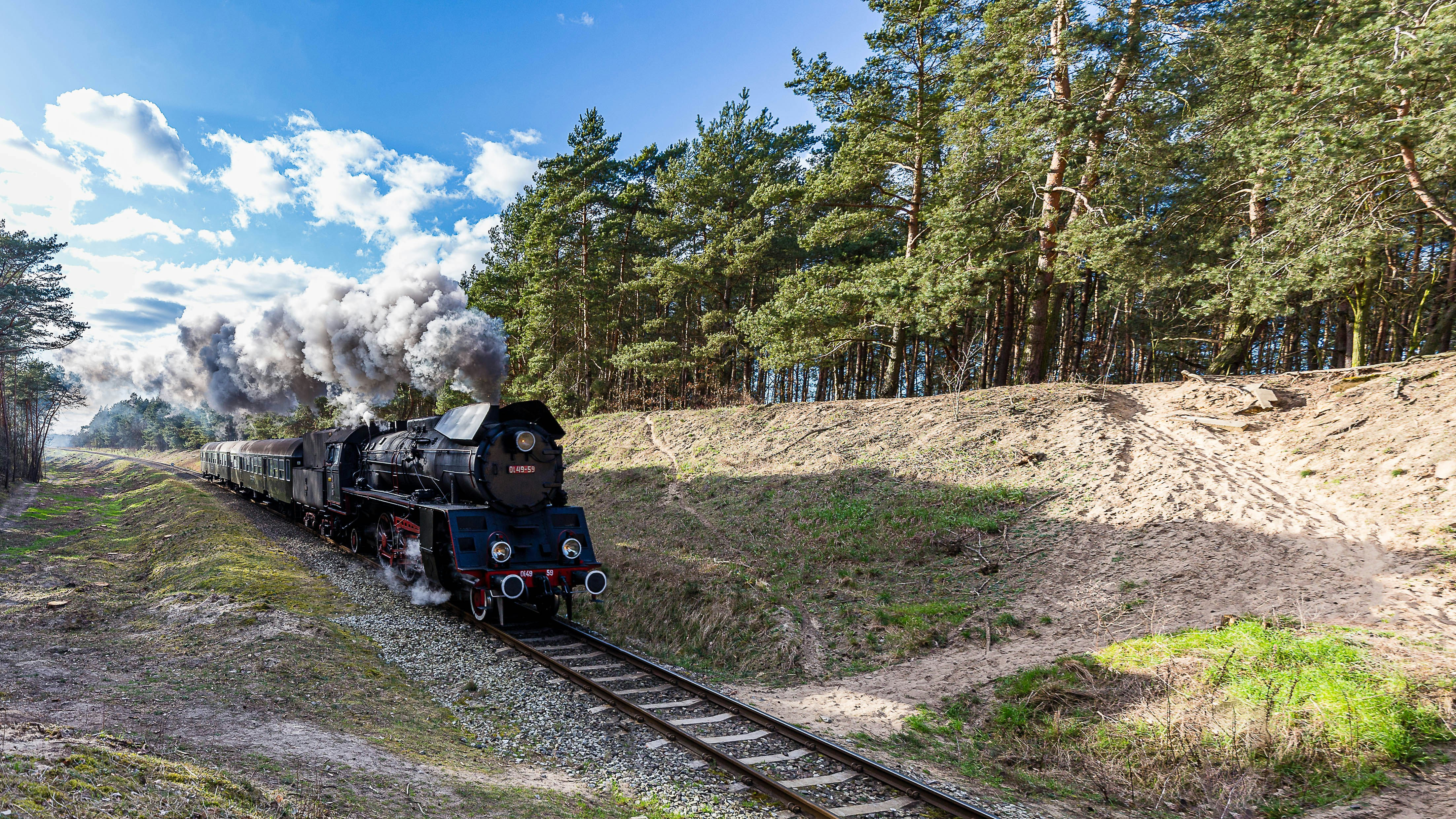 Vintage steam train traveling through a forested landscape under a bright blue sky with scattered clouds.