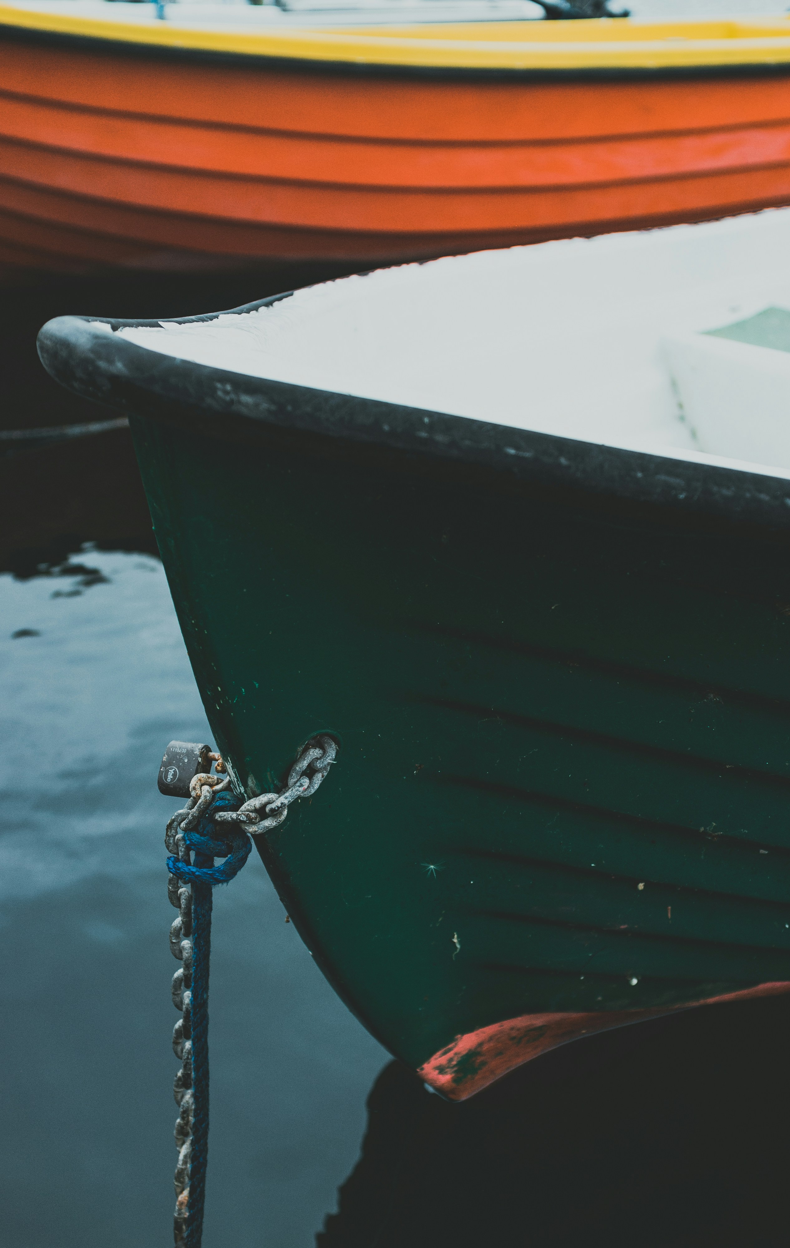 Close-up of a green boat's bow with a chain and rope, contrasted against a vibrant orange and yellow boat in the background. The calm water reflects the colors of the boats.