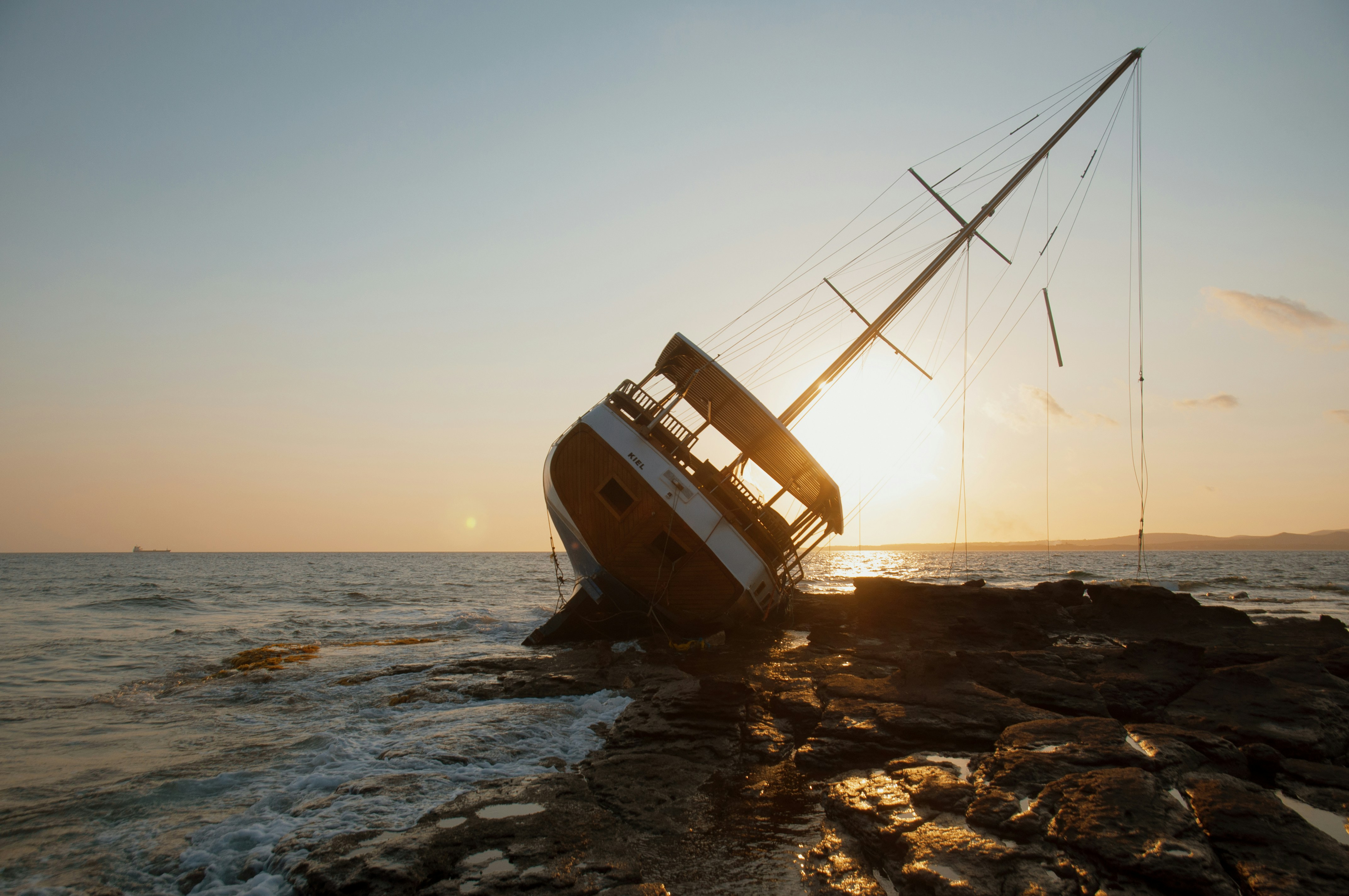 A beached ship tilts dramatically against the backdrop of a setting sun, highlighting its weathered hull and the rocky shoreline. The scene evokes a sense of nostalgia and abandonment.