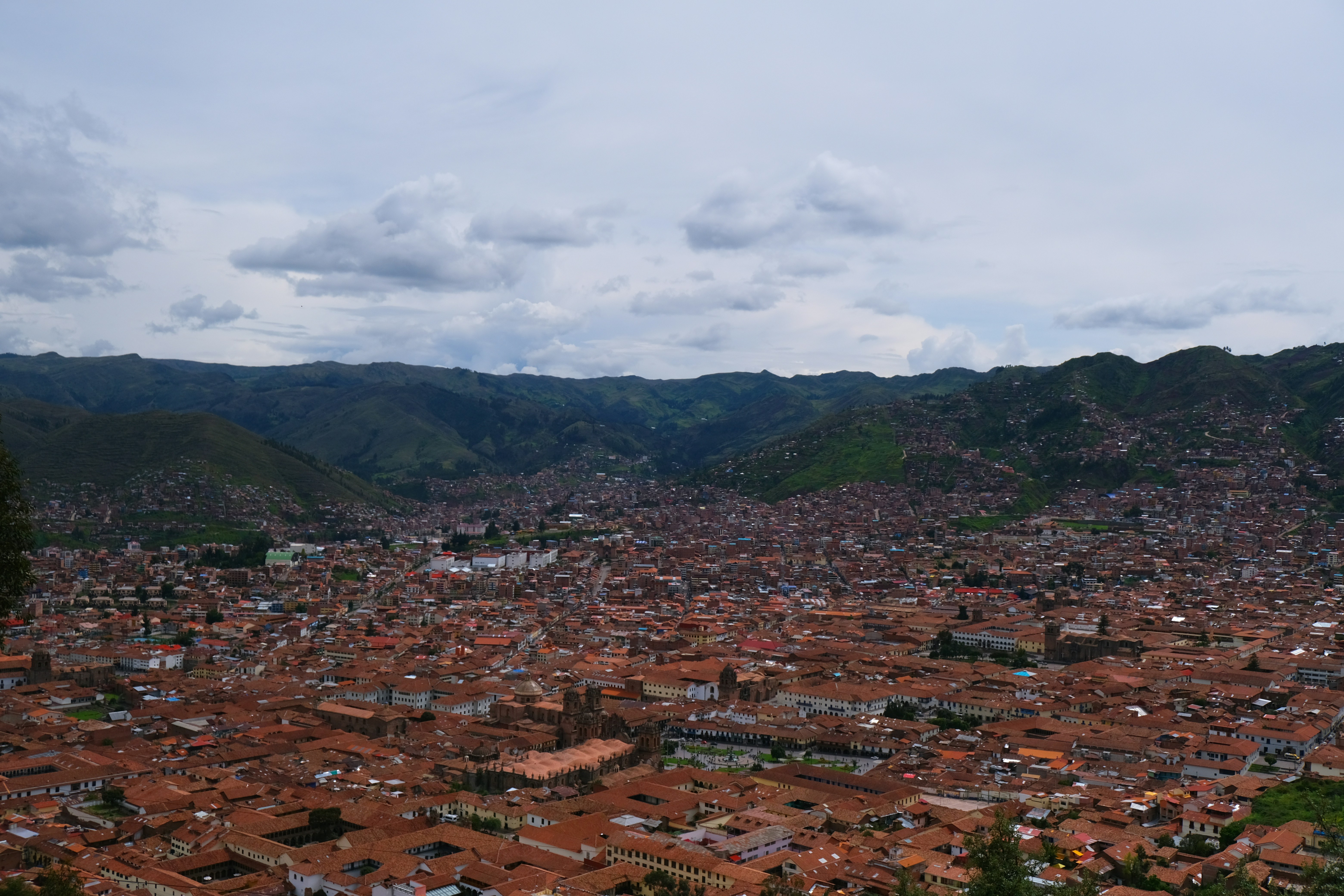 Expansive view of Cusco's terracotta rooftops nestled between lush green mountains under a cloudy sky.