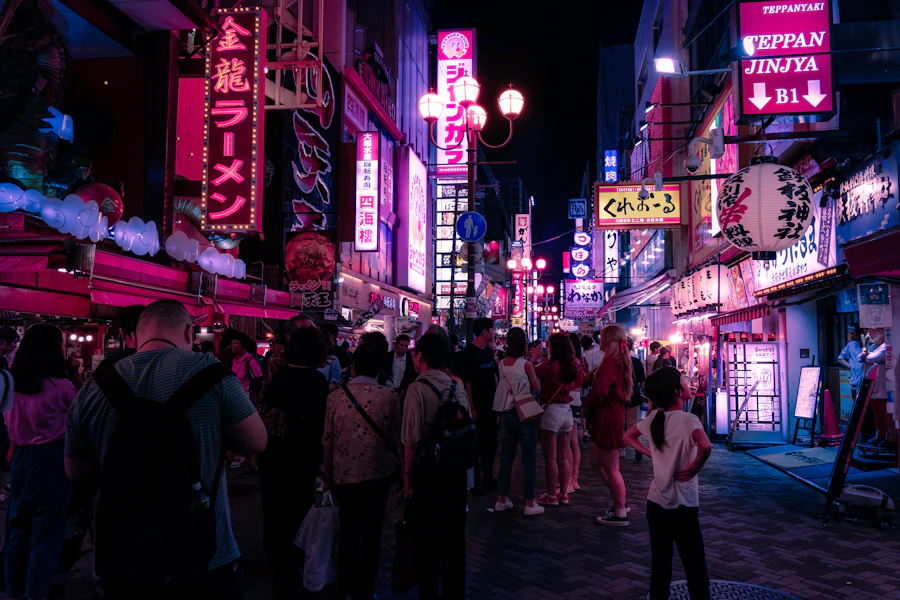Crowded Tokyo alley glowing with pink and blue neon signs, lanterns, and restaurant storefronts at night. People gather and walk through the lively street scene.