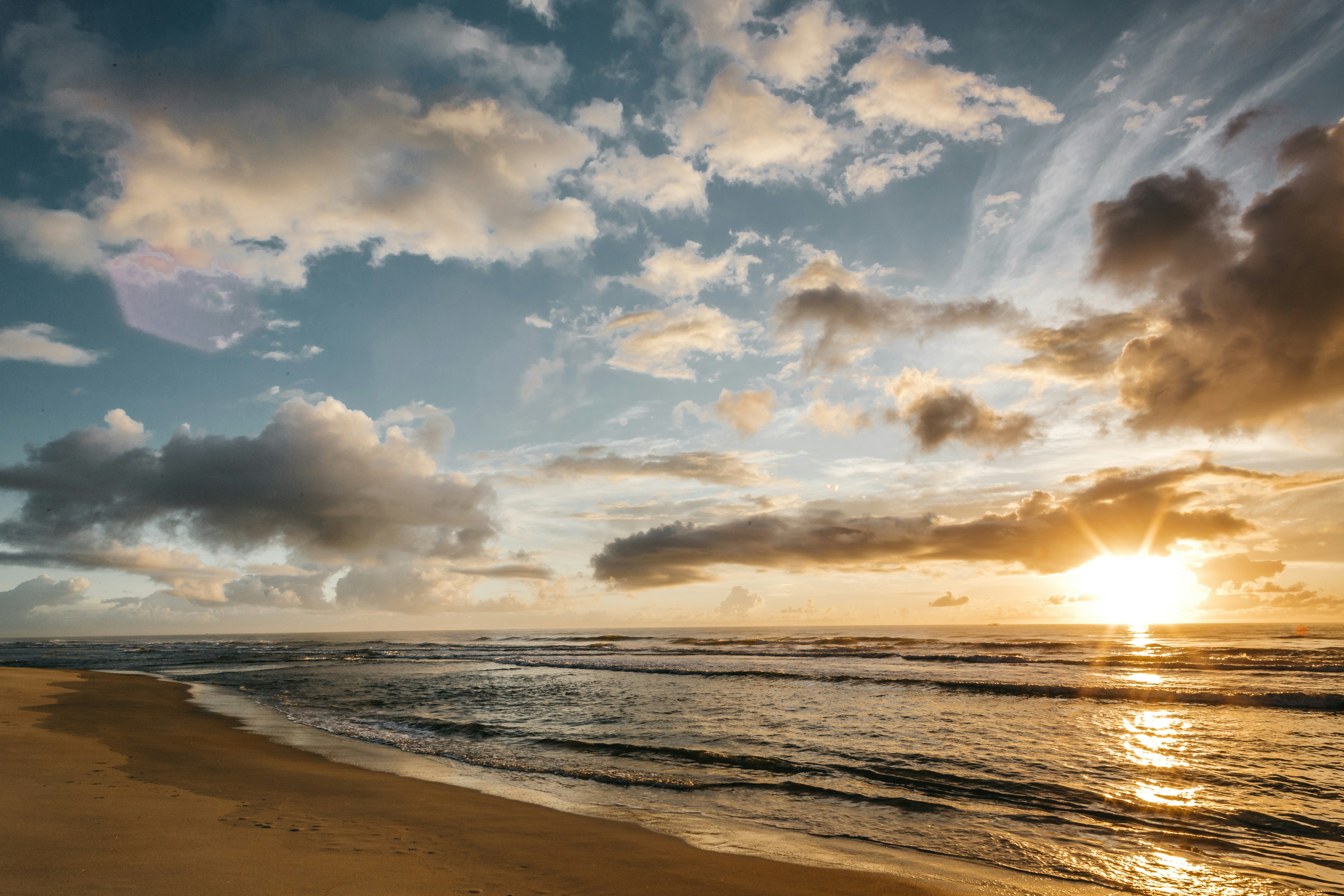 Golden sunrise casting reflections on a tranquil beach with scattered clouds in the sky.