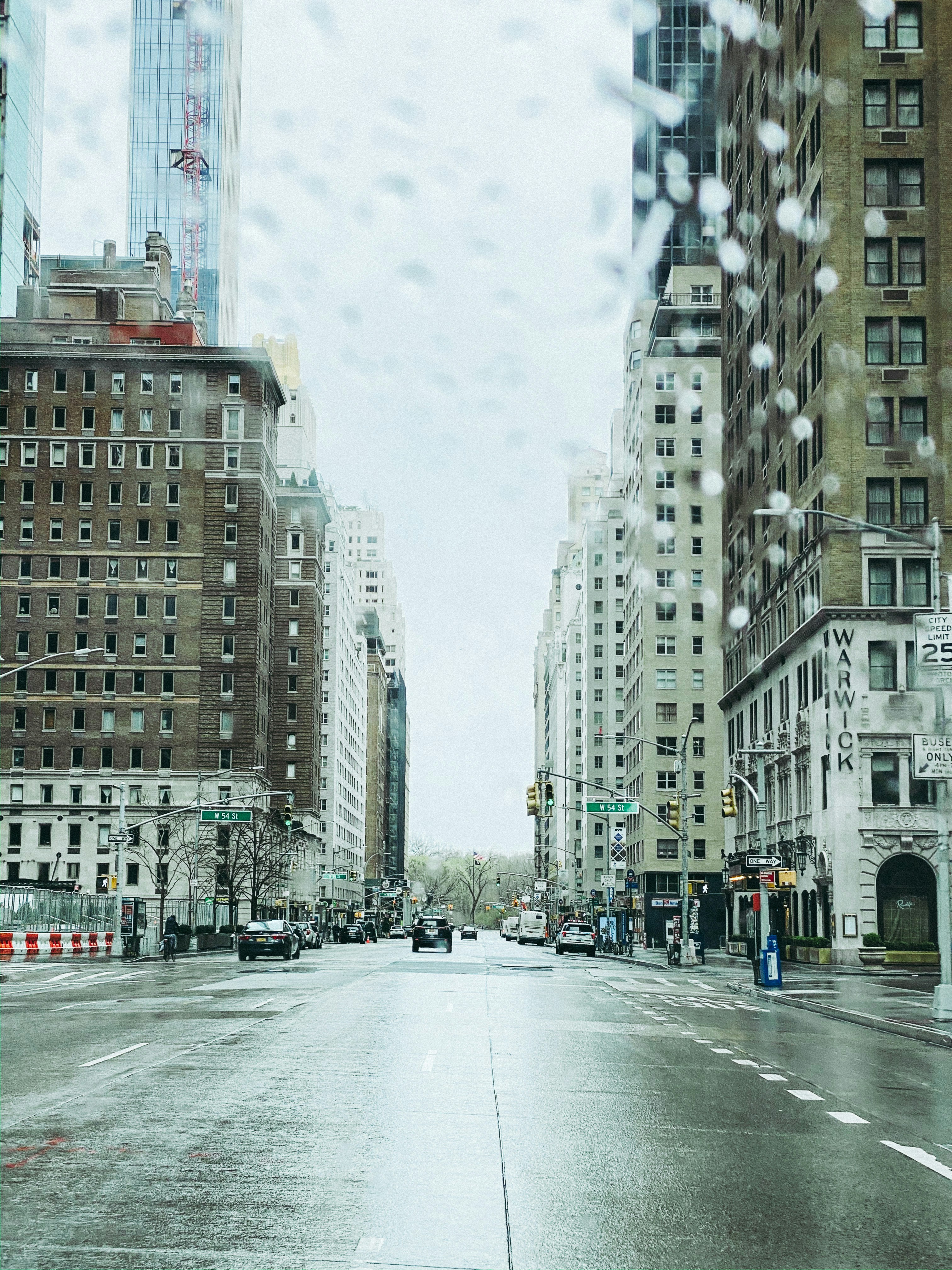 A rainy city street lined with tall buildings, reflecting the wet pavement and a muted sky. Traffic moves slowly under the gray atmosphere.