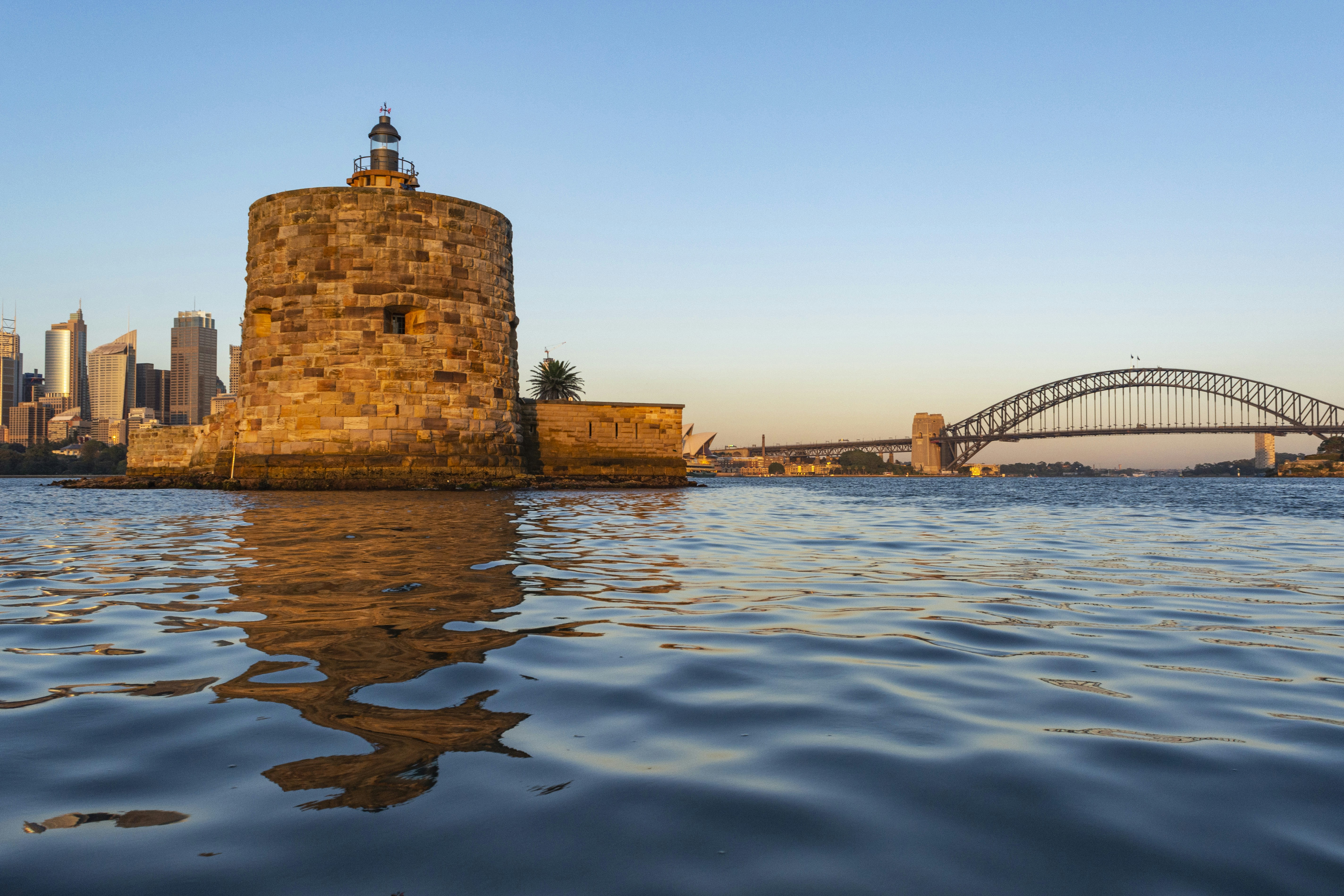 Historic stone fort reflecting in calm waters with a distant view of a city skyline and arched bridge at sunset.