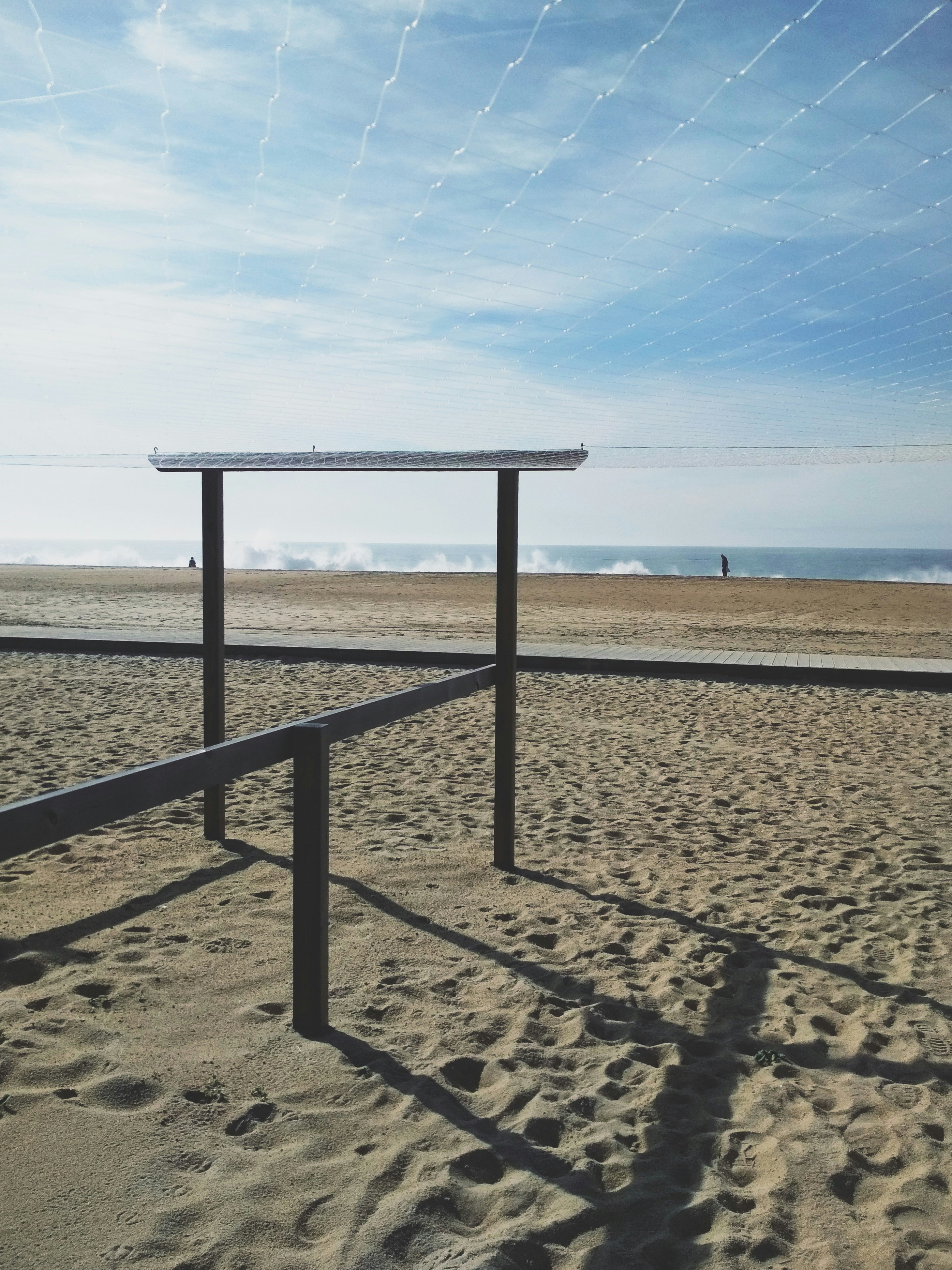 Minimalist beach scene showing a metal railing casting long shadows on sand beneath a grid of overhead netting, drawing the eye toward the distant horizon.