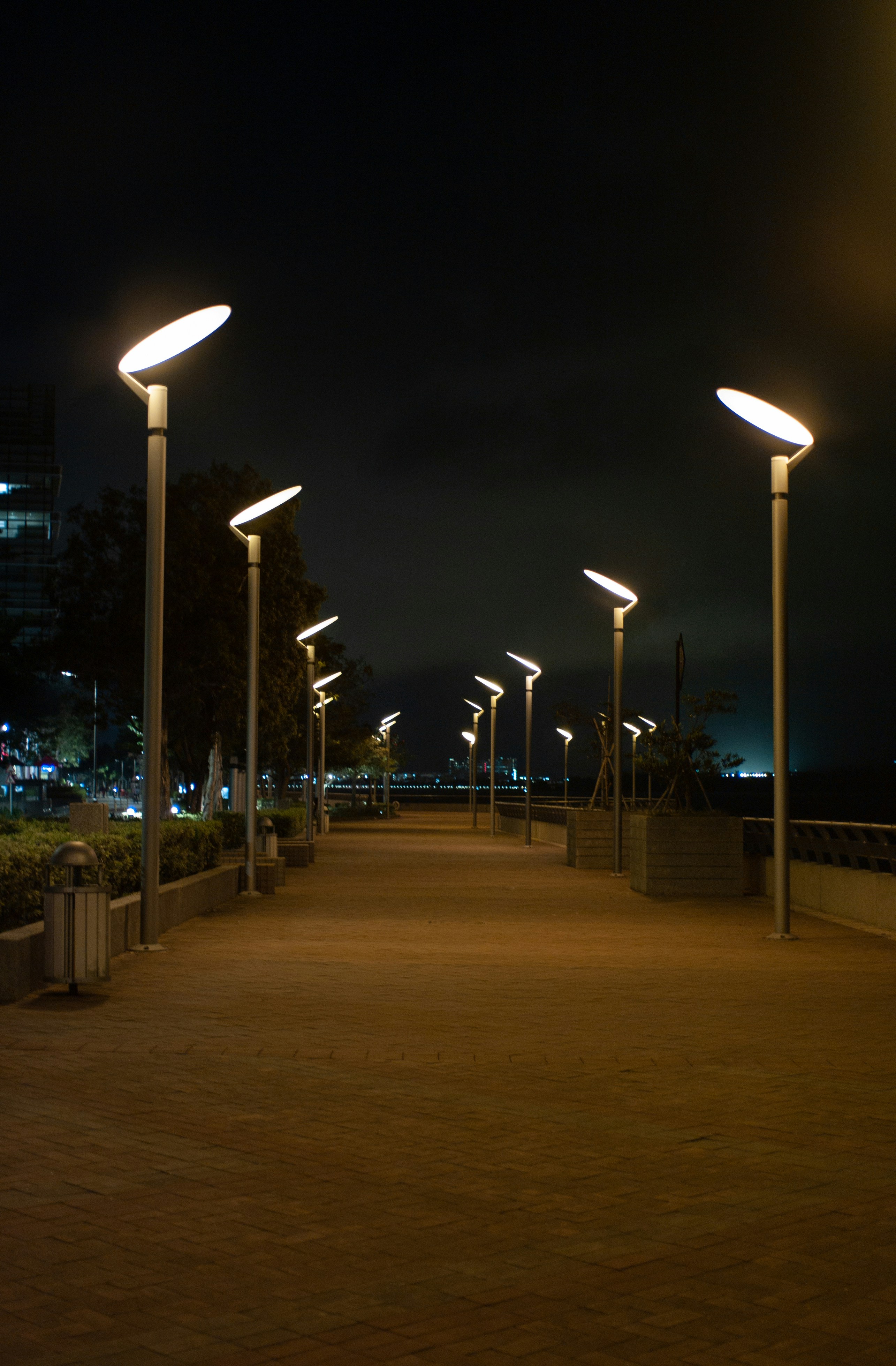 A serene walkway lined with modern street lamps, casting soft light against the dark night sky. The scene invites a peaceful evening stroll.