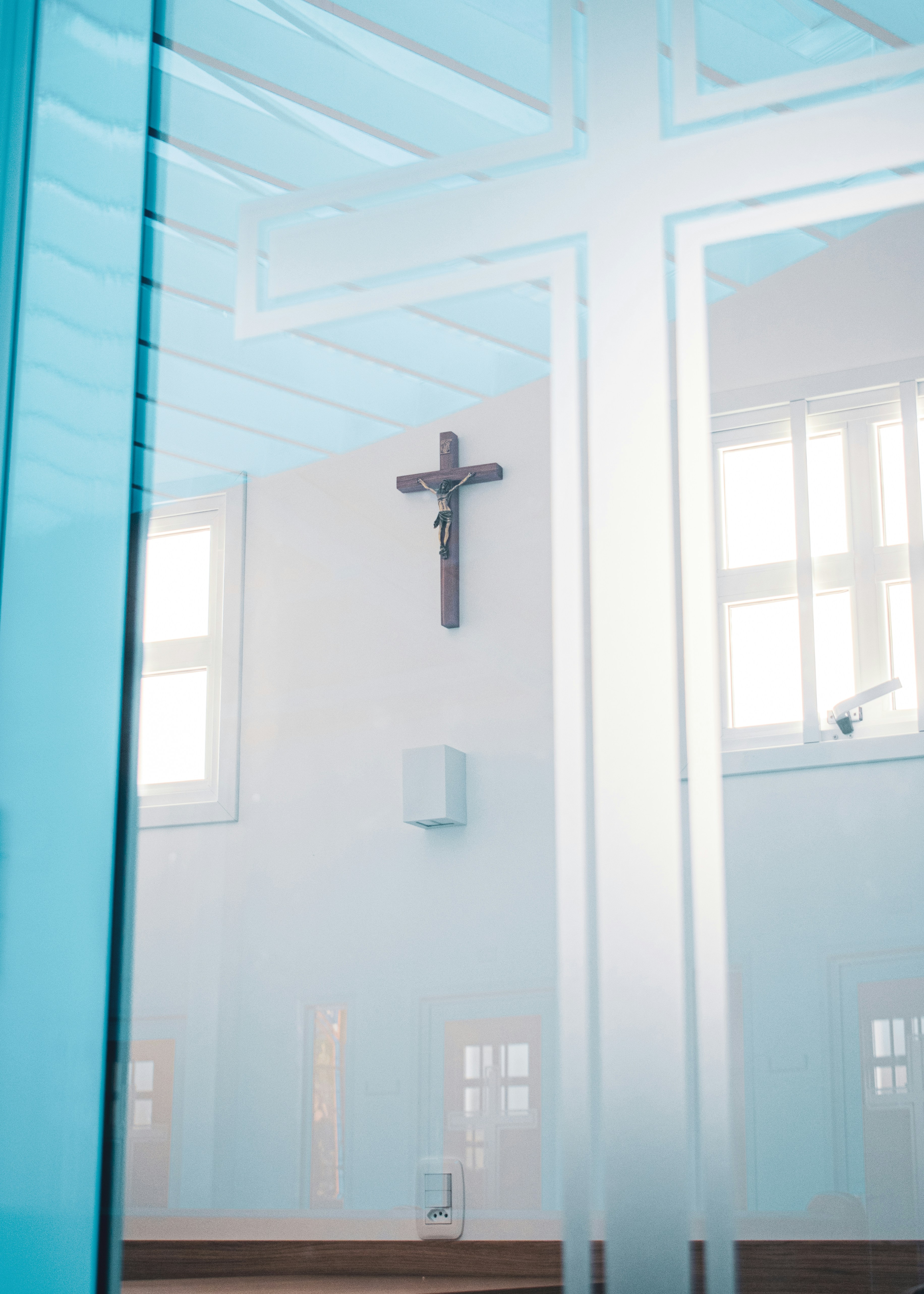 Cross reflected in a blue-tinted glass door with bright windows in the background.