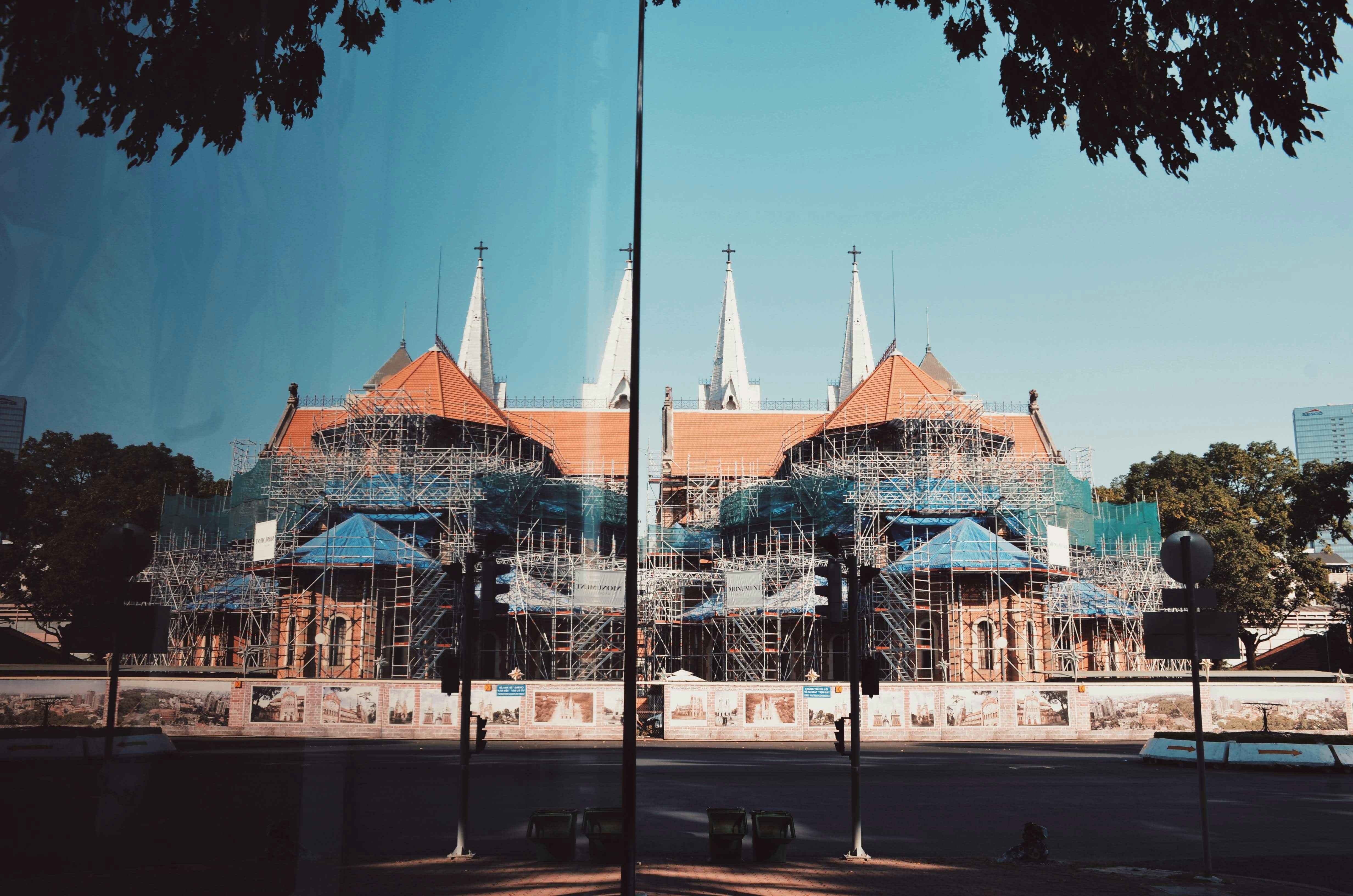 Historic building with pointed towers and scaffolding, reflected symmetrically in glass under a clear blue sky.