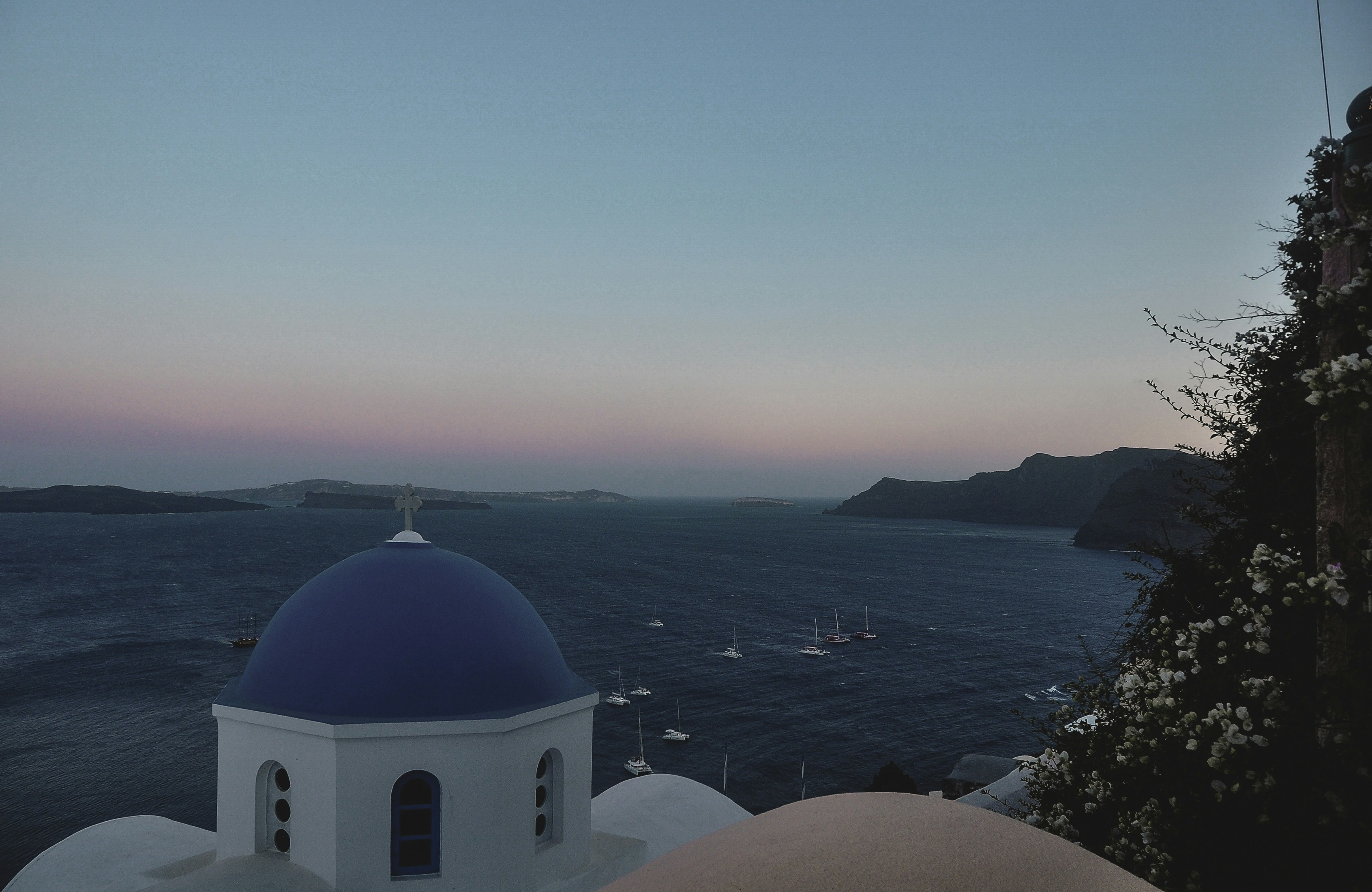 White-domed church overlooking the Aegean Sea at dusk with sailboats dotting the horizon.