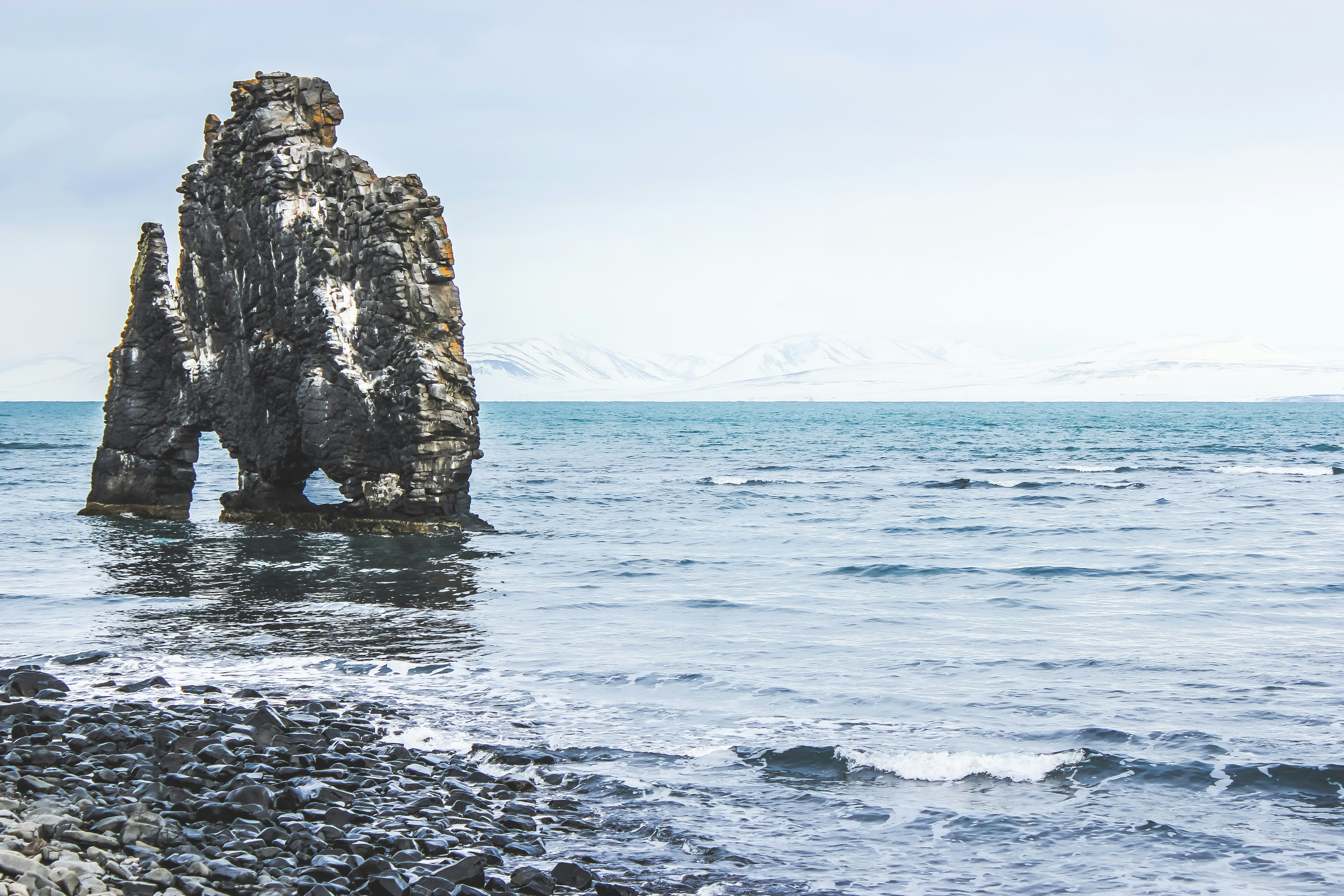 A rugged rock formation stands tall amidst the tranquil sea, with distant snow-capped mountains creating a serene backdrop.
