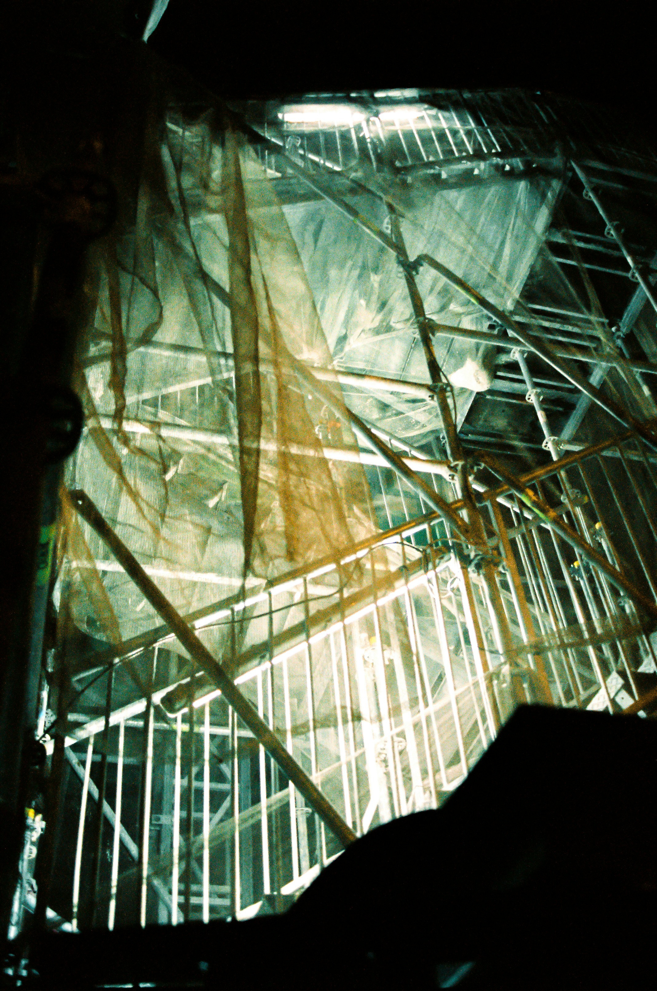 Abstract view of scaffolding illuminated by artificial light, revealing layers of fabric and metal. The composition creates a sense of depth and intrigue.