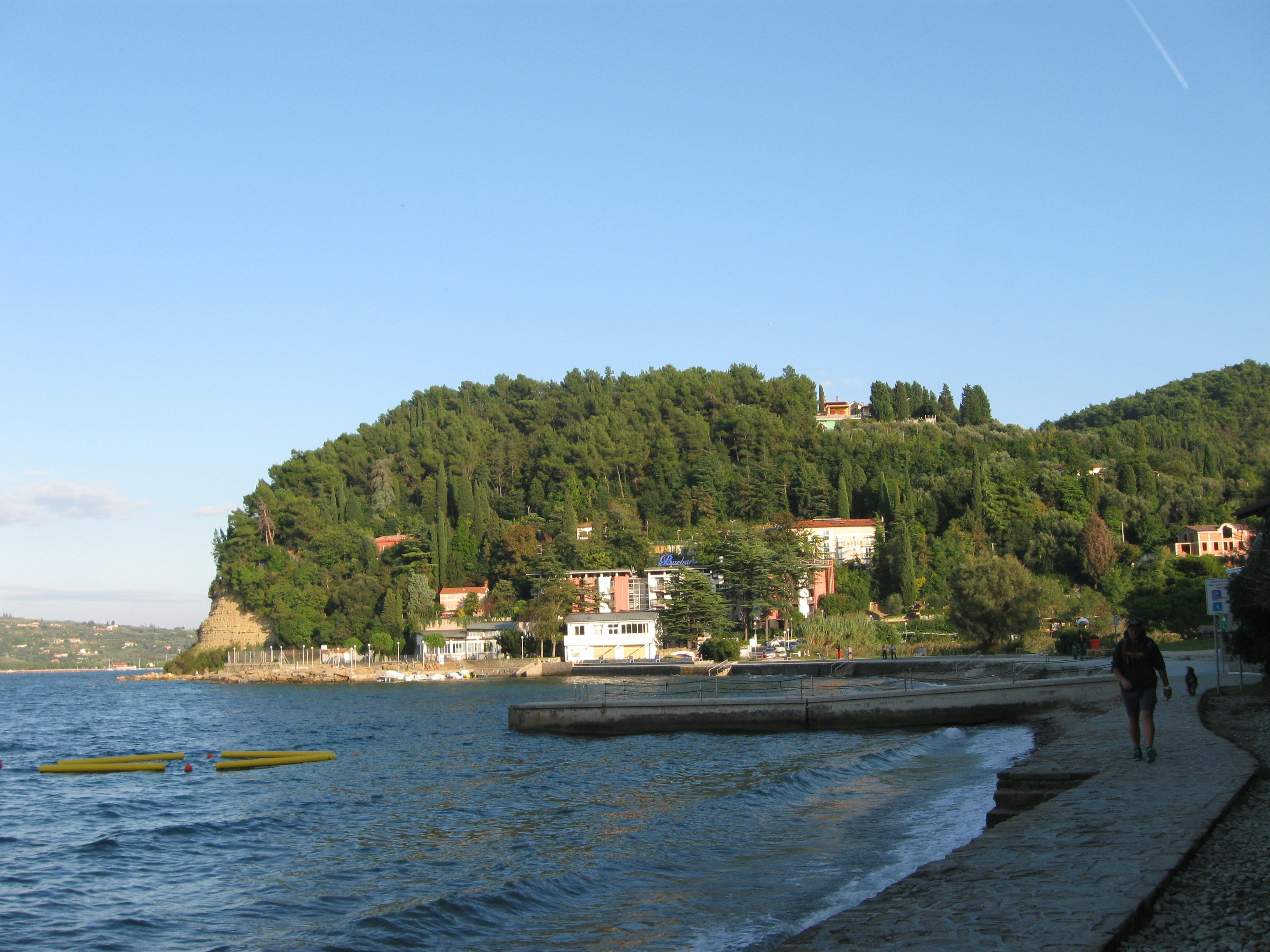 Coastal promenade along a pebble path beside a calm cove, backed by a forested hillside and white houses. Two yellow paddle boards drift near the water while a lone figure walks the path.