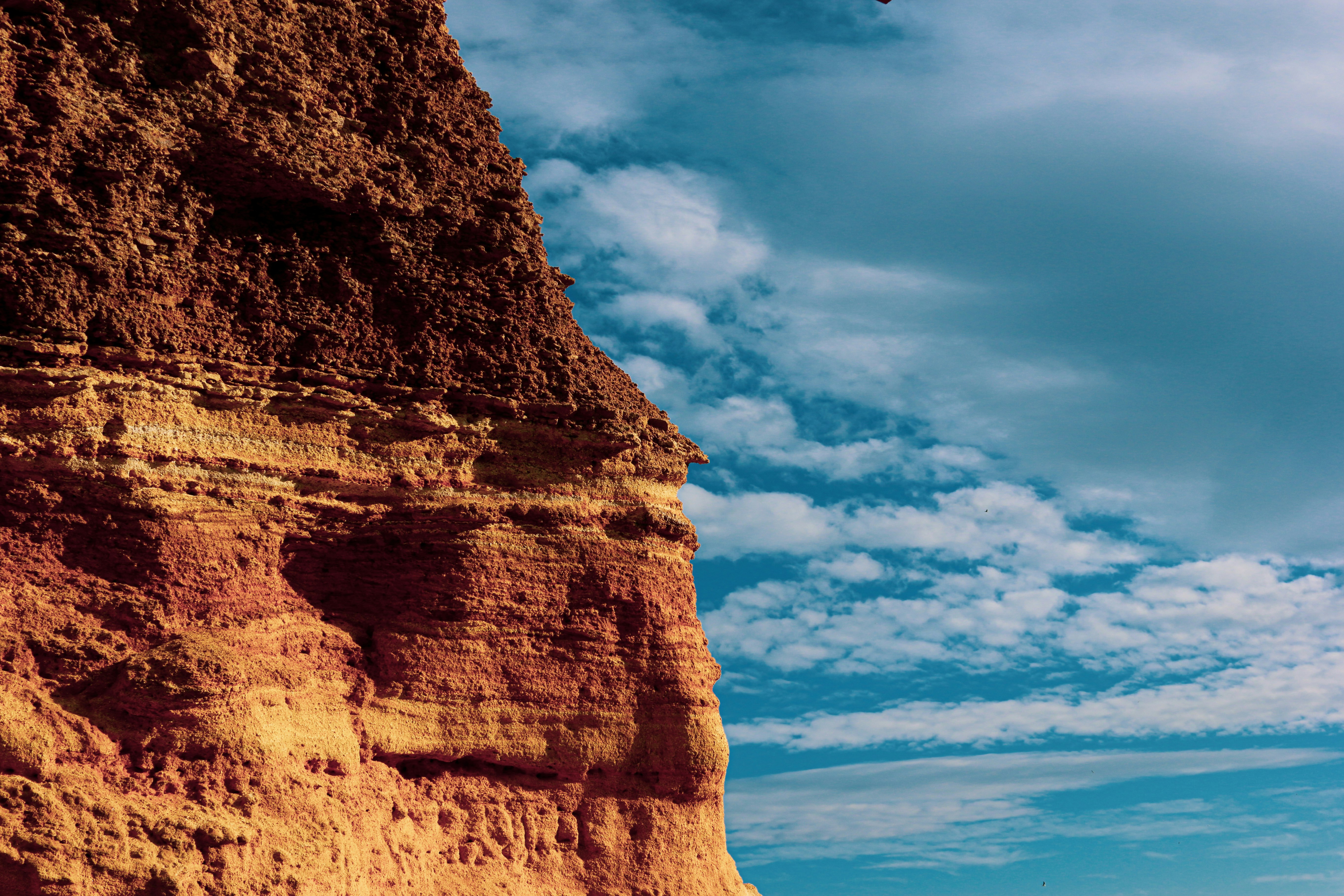 Steep rocky cliff with layered textures set against a vibrant blue sky scattered with clouds.