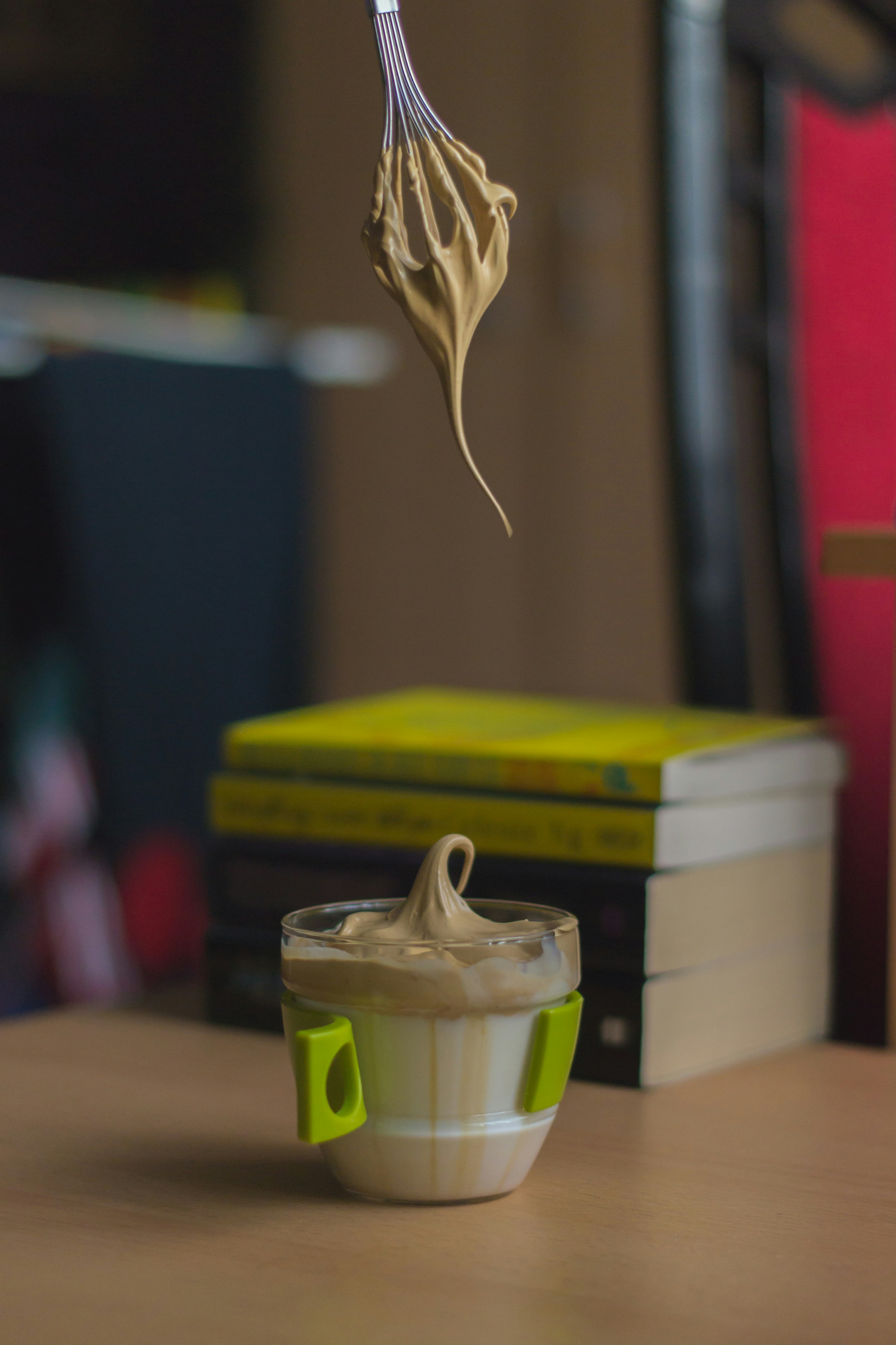 Creamy swirl of chocolate mousse hanging above a mug, with a stack of books blurred in the background. The scene emphasizes a soft, vertical motion and a warm, cozy kitchen vibe.