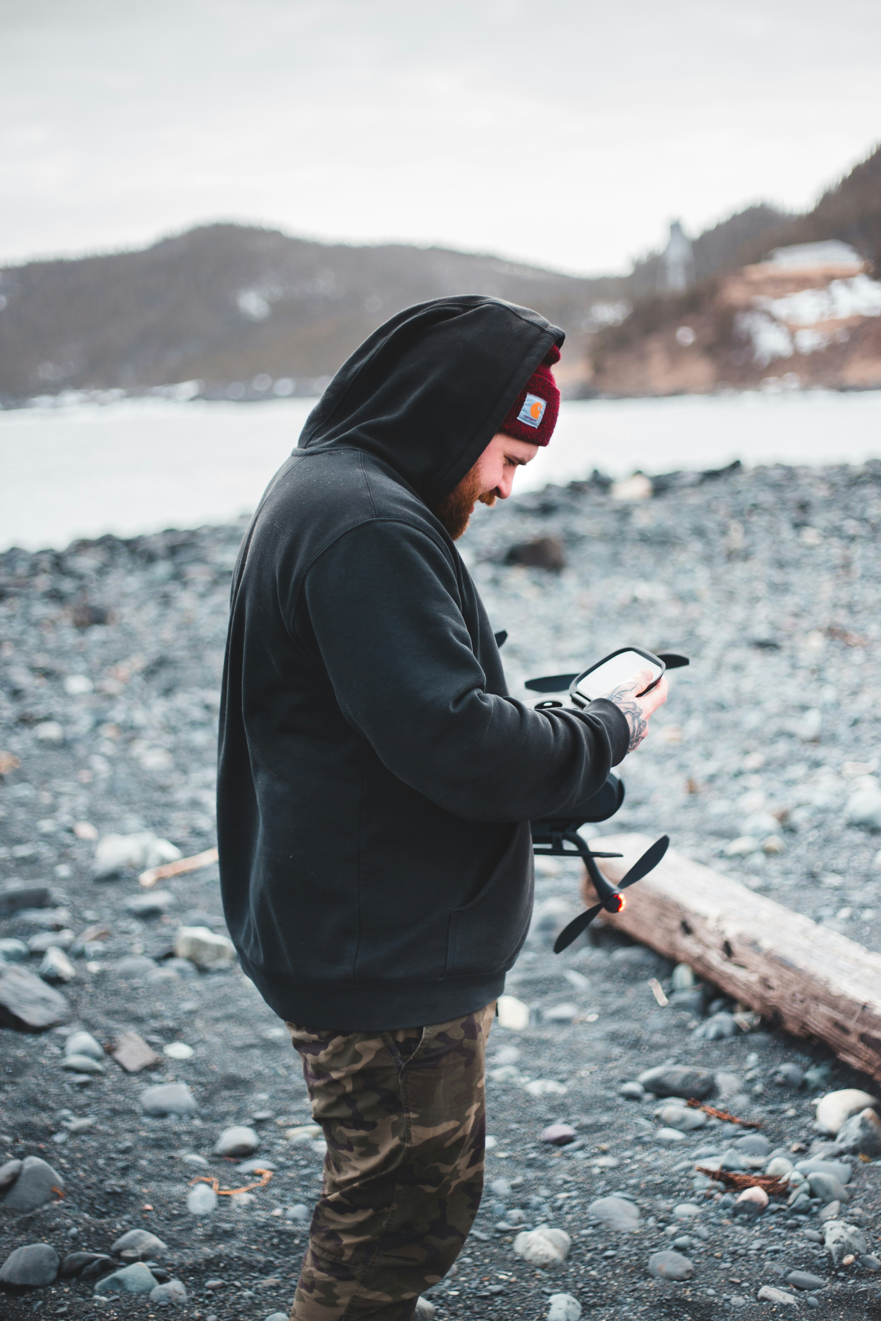 A Quiet Moment with the Compass: Man in Hoodie Engaged with a Pocket Device on a Rocky Shore