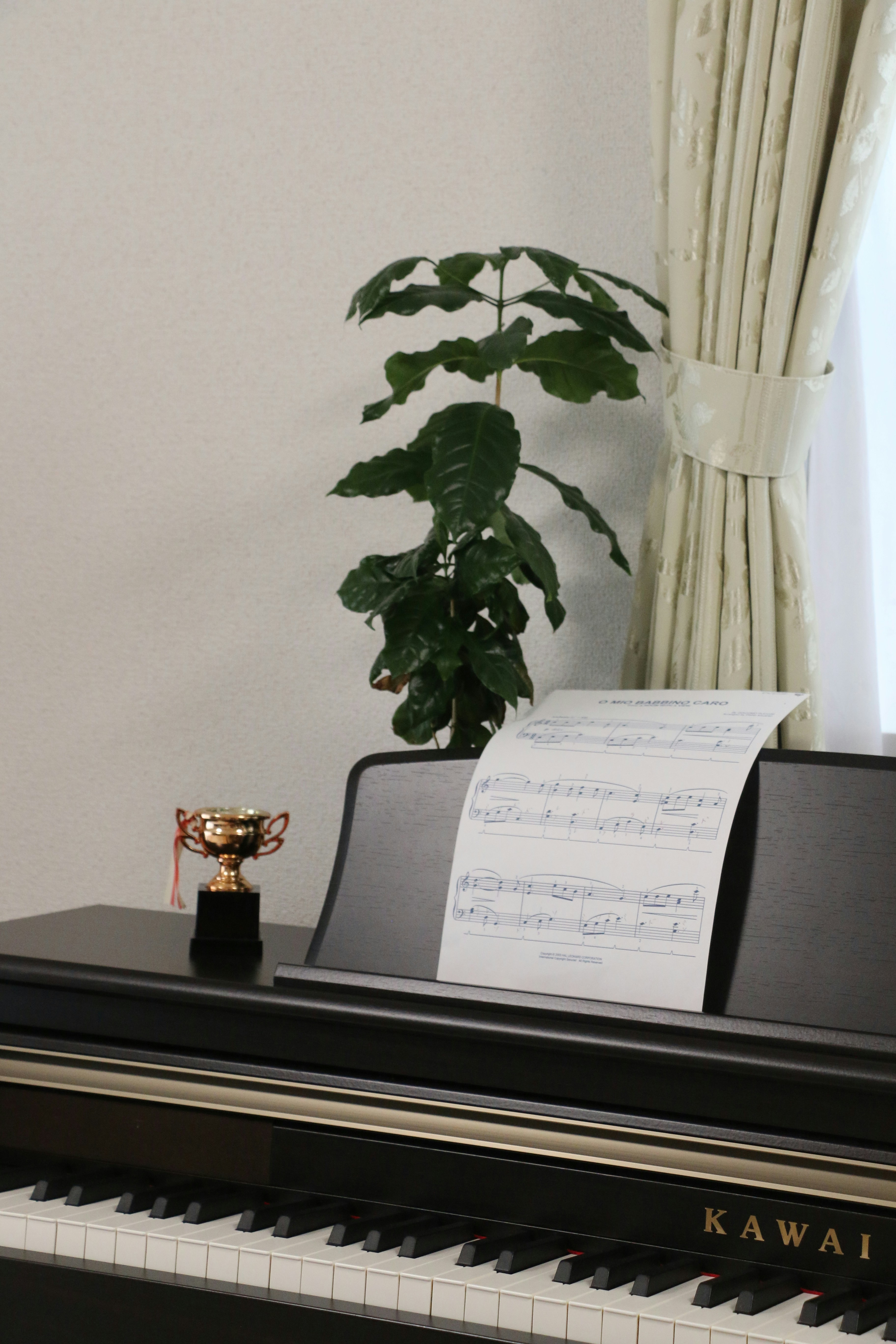 Quiet Interlude: A Kawai Piano, Sheet Music, and a Glittering Trophy in Soft Window Light