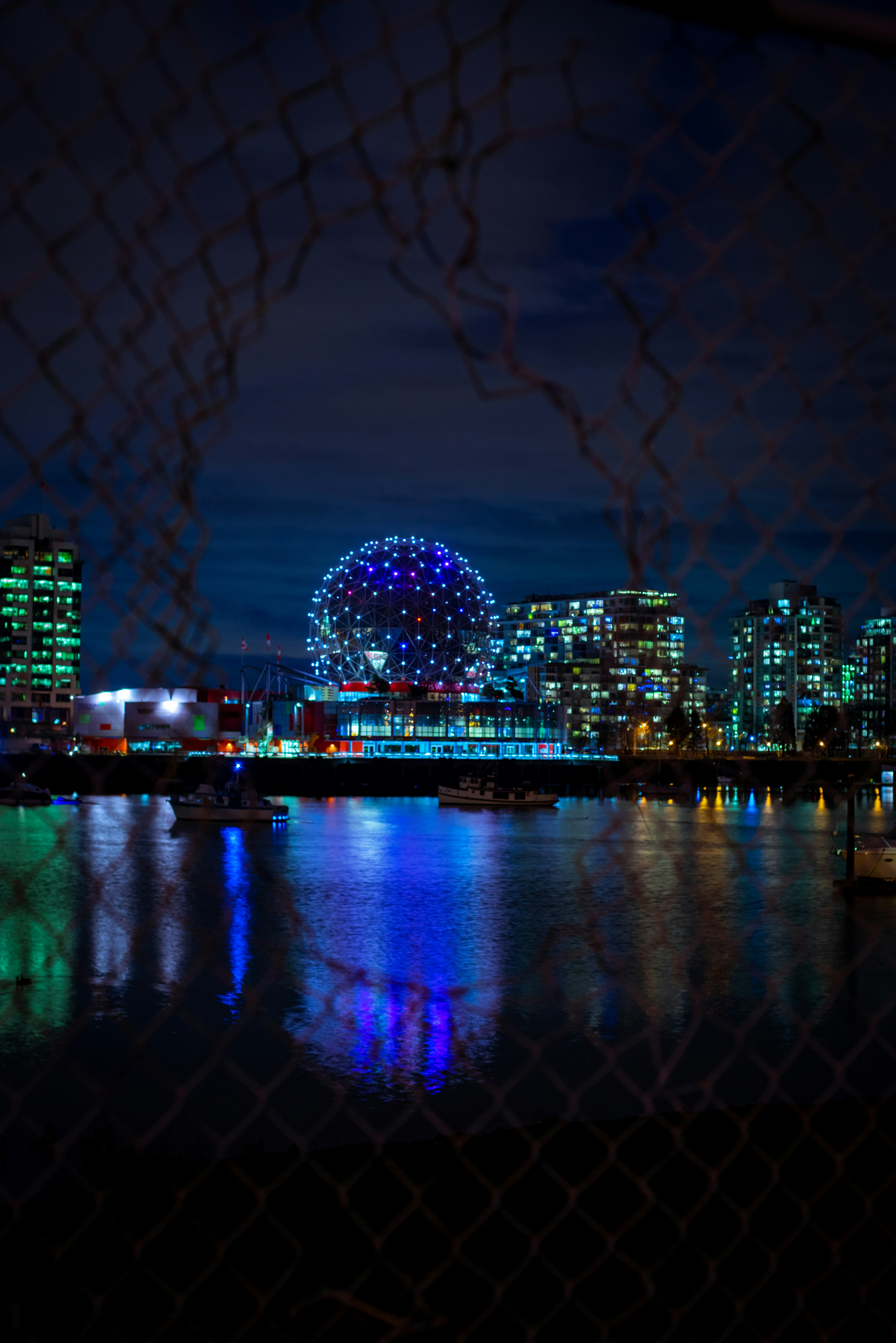 Neon Globe Over the Marina: Night Reflections Through a Chain-Link Veil