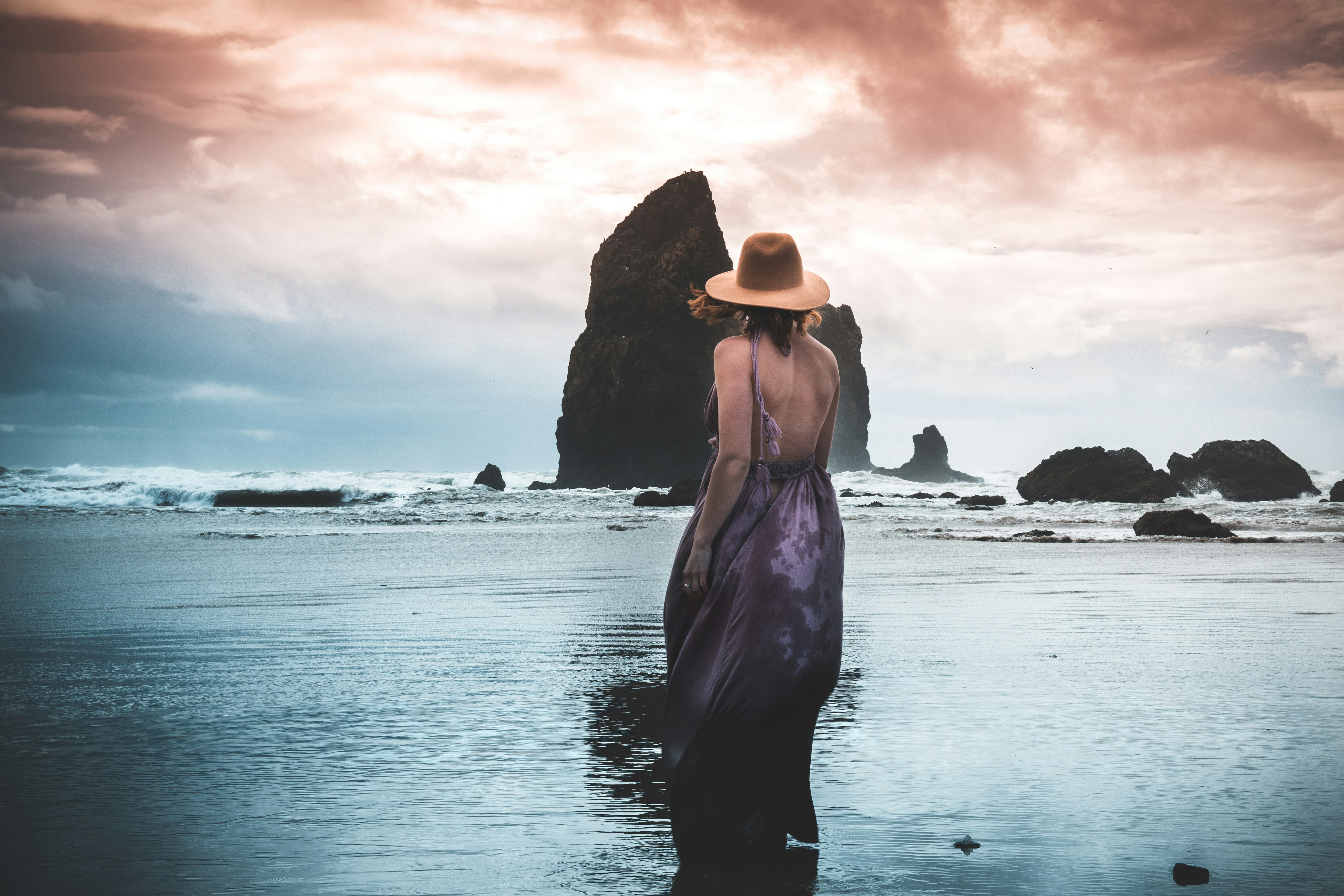 Woman in blue dress and hat walking towards sea stacks on a tranquil beach at dusk.