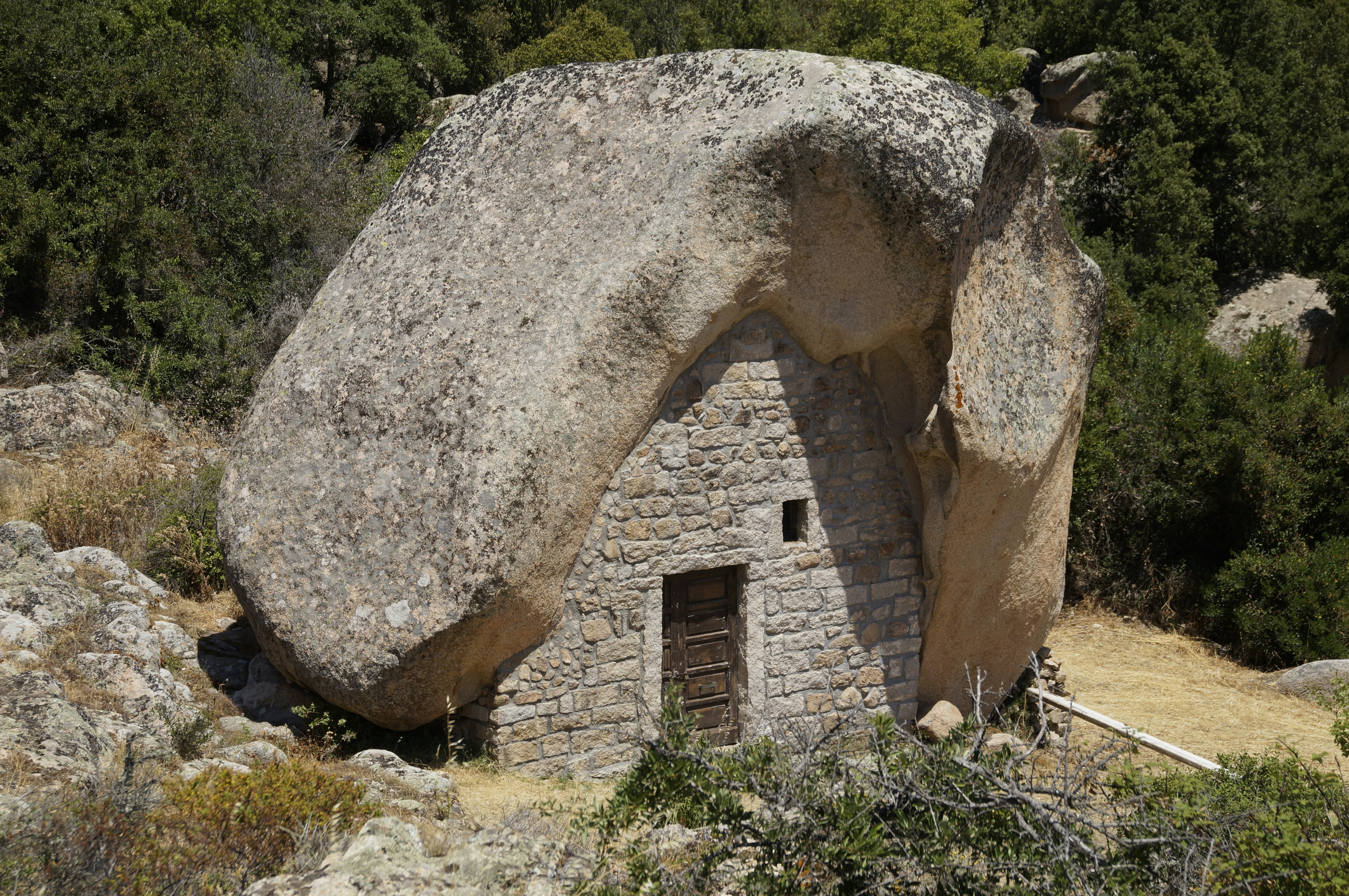 A small stone structure nestled beneath a massive boulder, surrounded by lush greenery. The scene evokes a sense of harmony between nature and architecture.