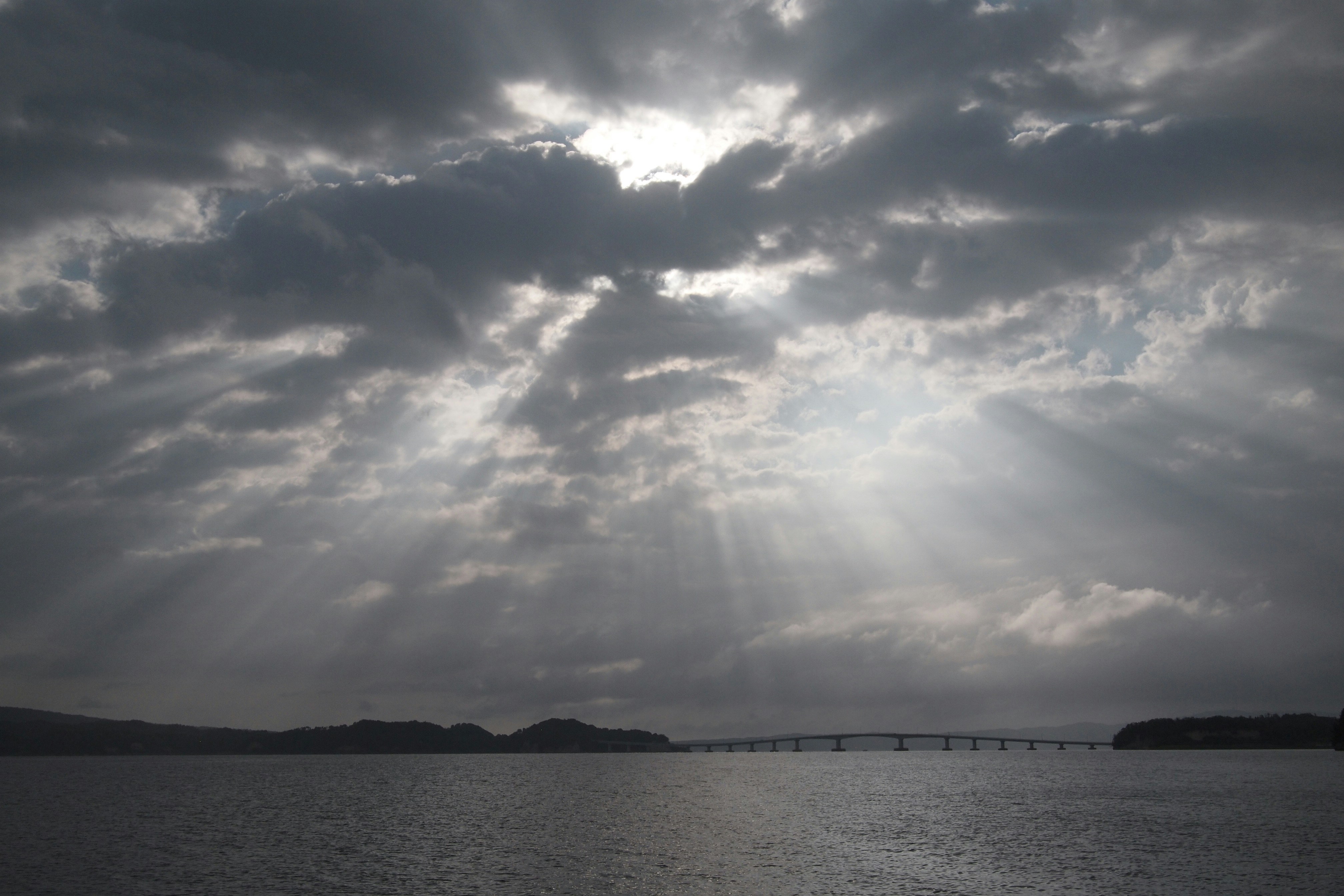 Sunlight breaks through dark clouds, casting radiant beams over a calm sea with a distant bridge. The scene evokes a sense of tranquility and wonder.