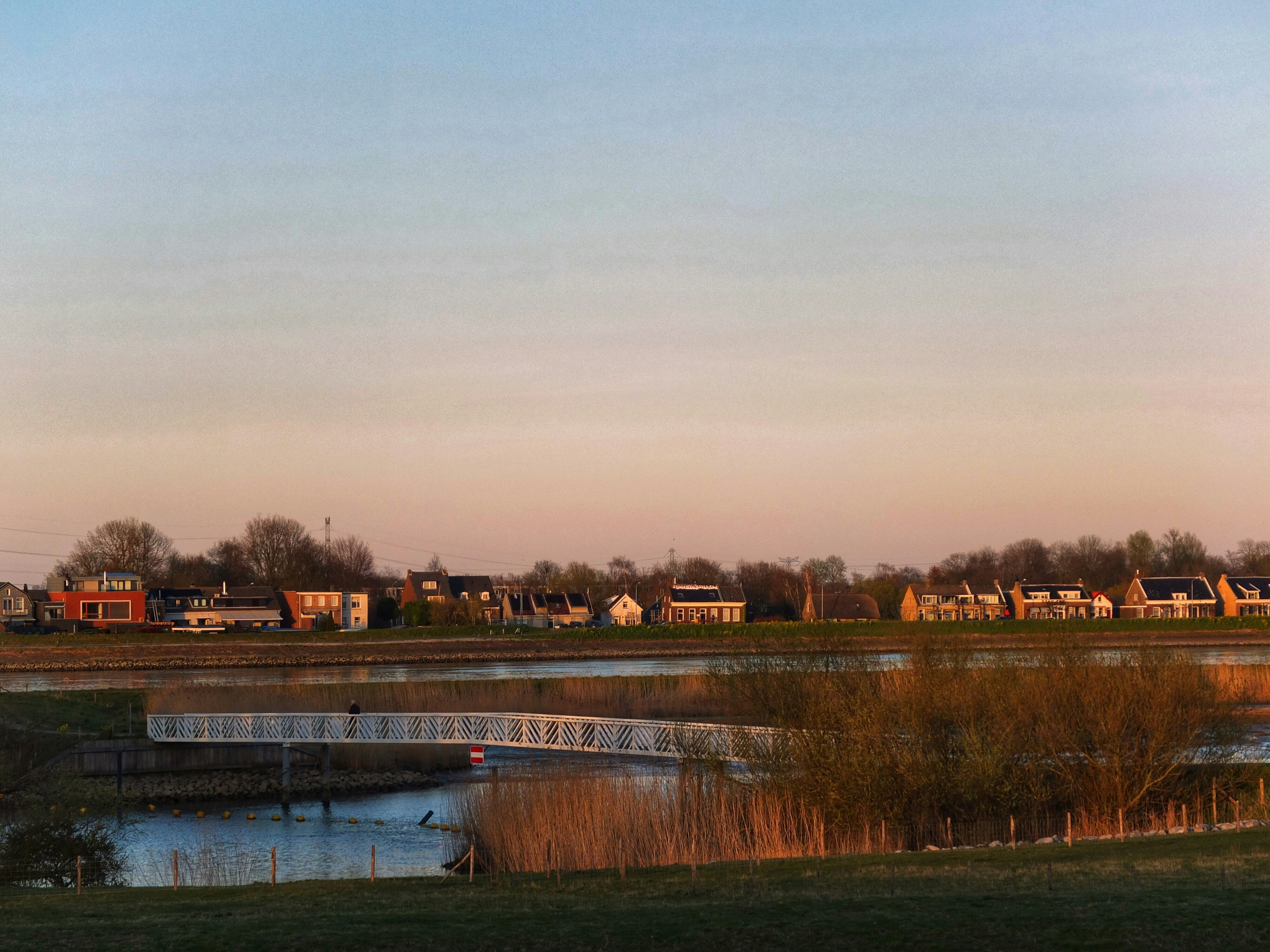 Charming houses line the riverbank, reflecting the soft hues of dusk, with a quaint wooden bridge crossing the water. 