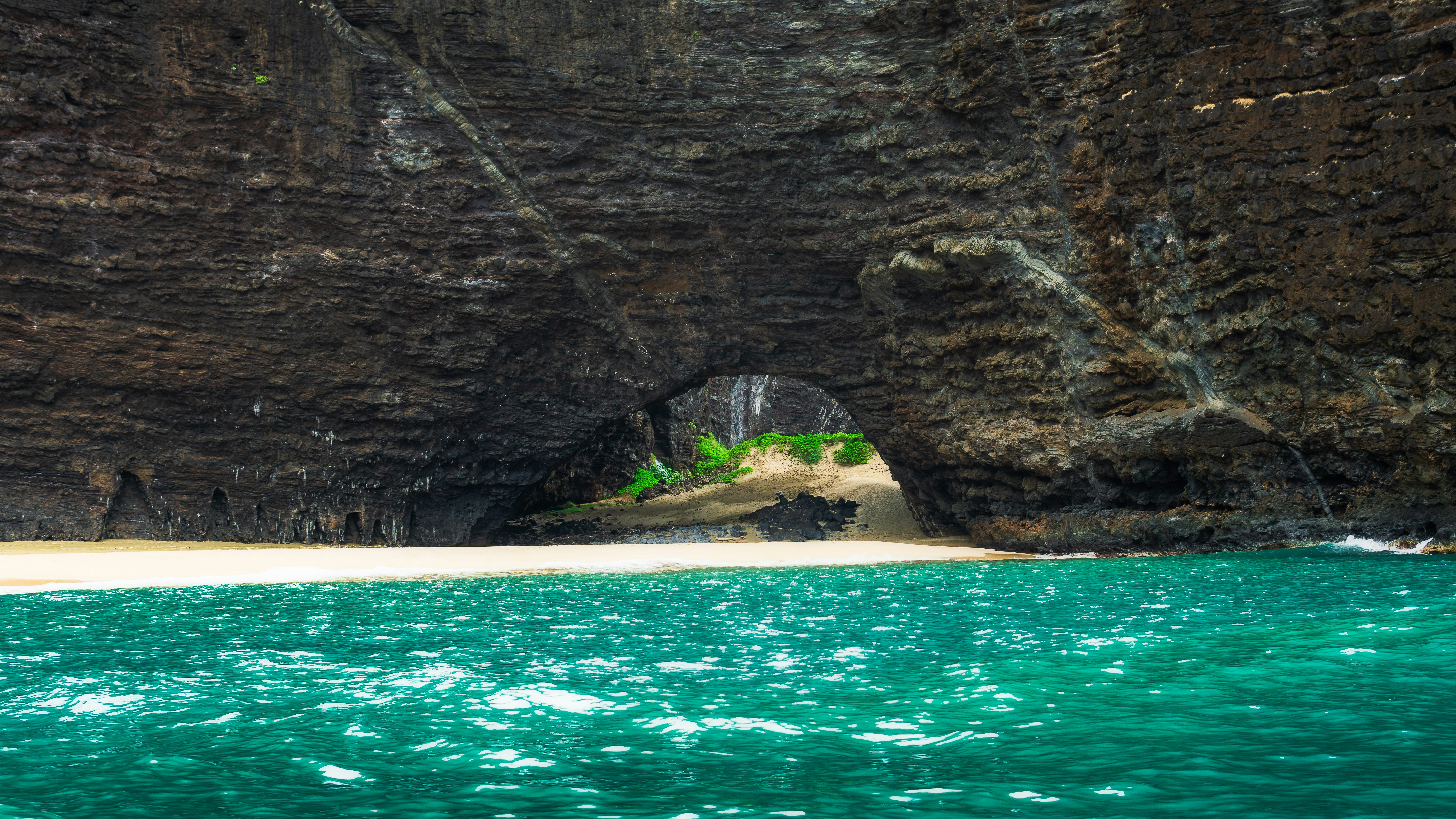 Turquoise ocean waves meet a sandy shore beneath a natural rock arch on Kauai's Na Pali Coast.