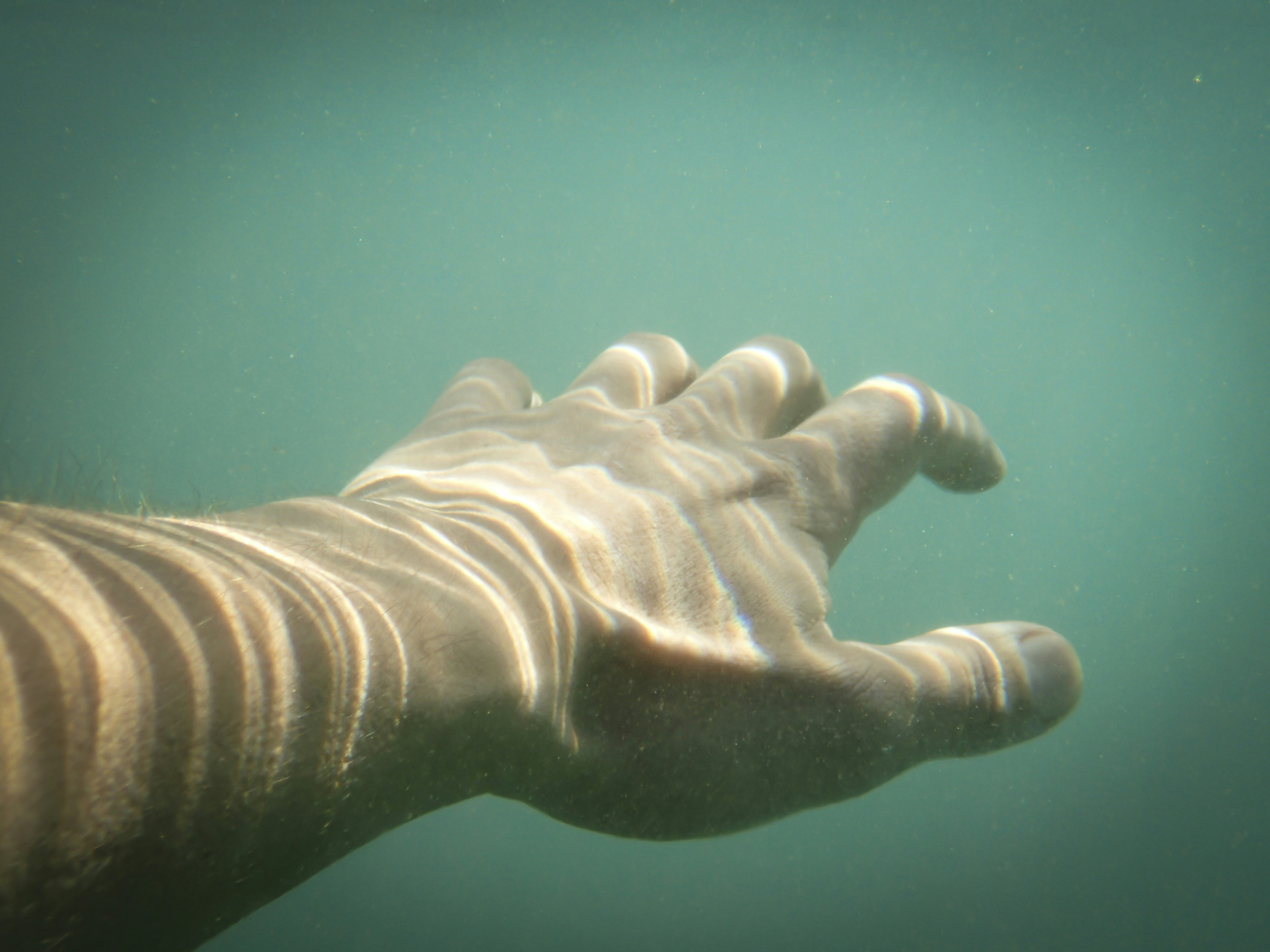 A submerged hand reaches out, creating ripples in the water while sunlight filters through the surface. The play of light and shadow adds depth to the scene.