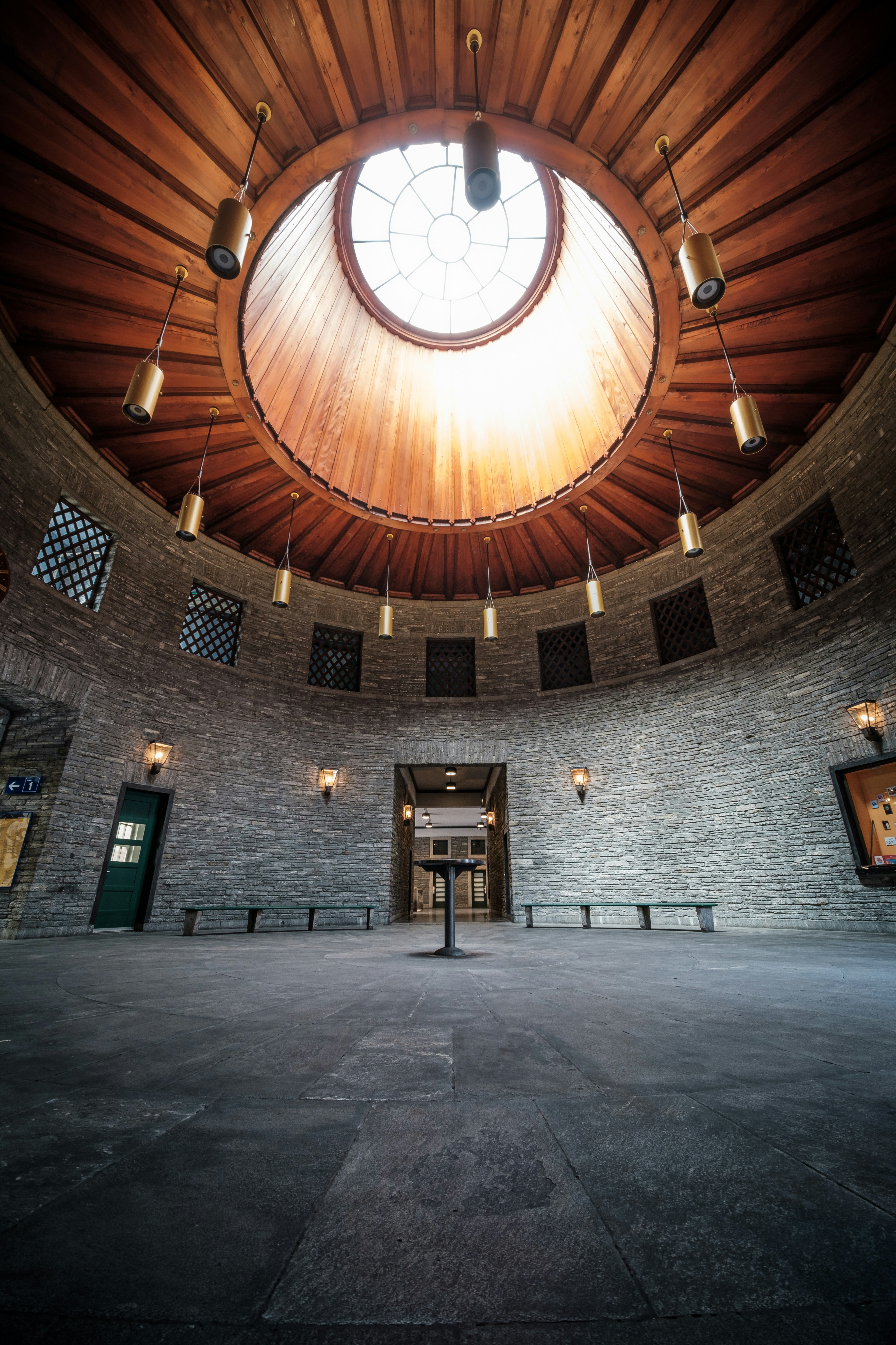 Circular interior space featuring a wooden dome and stone walls, illuminated by hanging lights. The design emphasizes symmetry and architectural elegance.