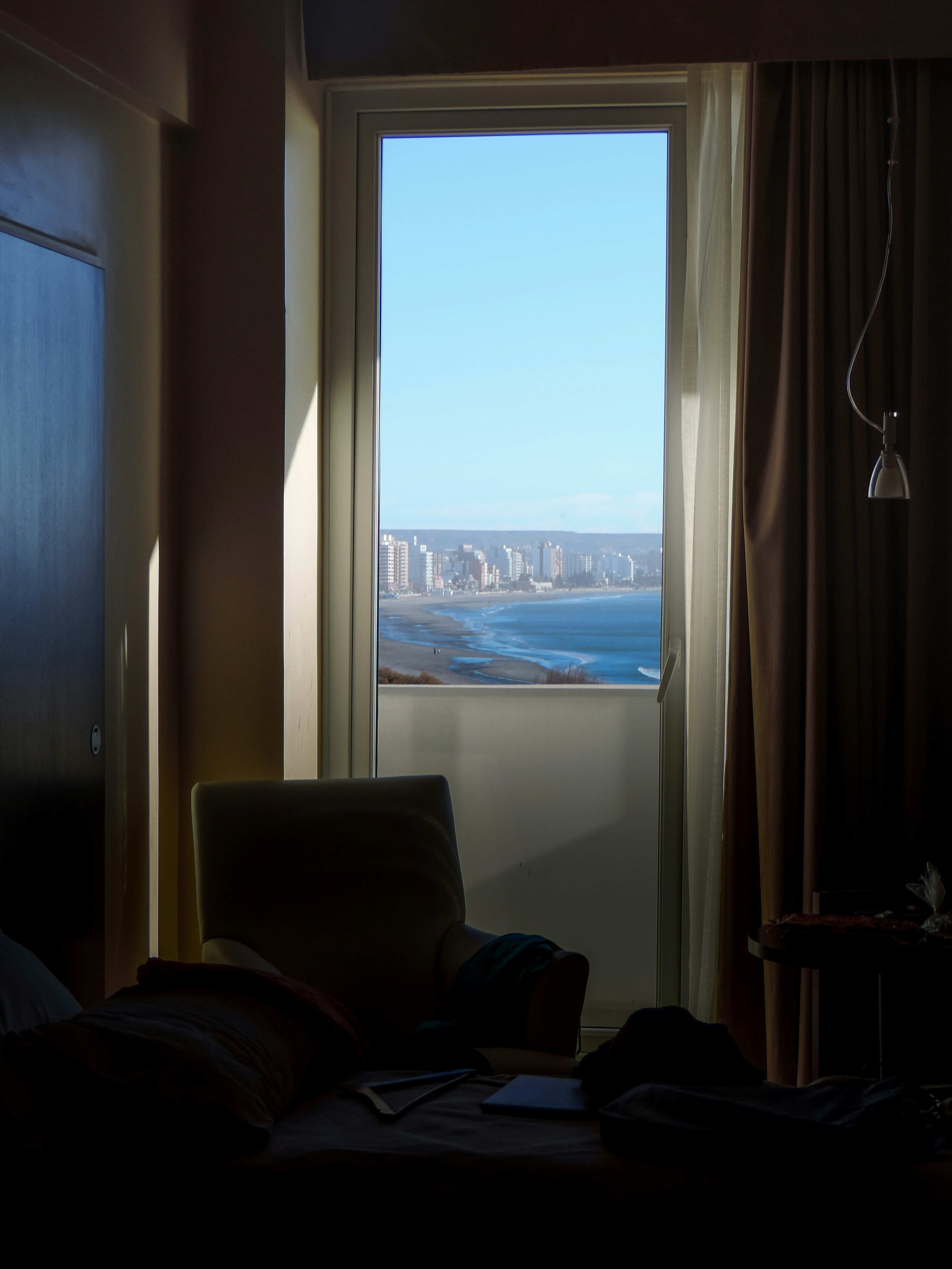 Warm hotel room with a window view of the sea and distant buildings, balcony beyond the glass; foreground shows a chair and dimly lit seating area.
