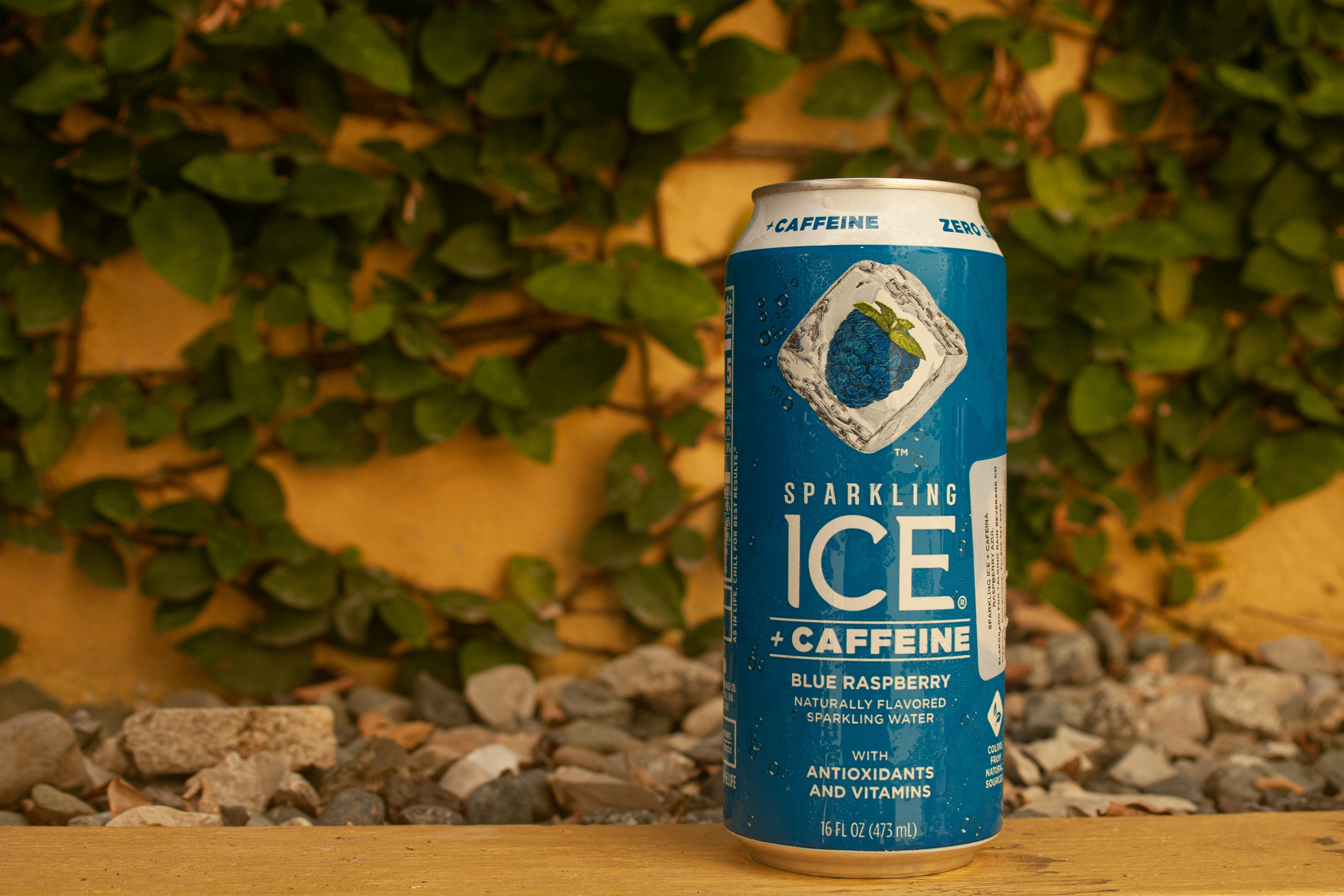 Blue Raspberry Sparkling ICE can sits on a rocky ledge in front of a sunlit yellow wall covered with green ivy.